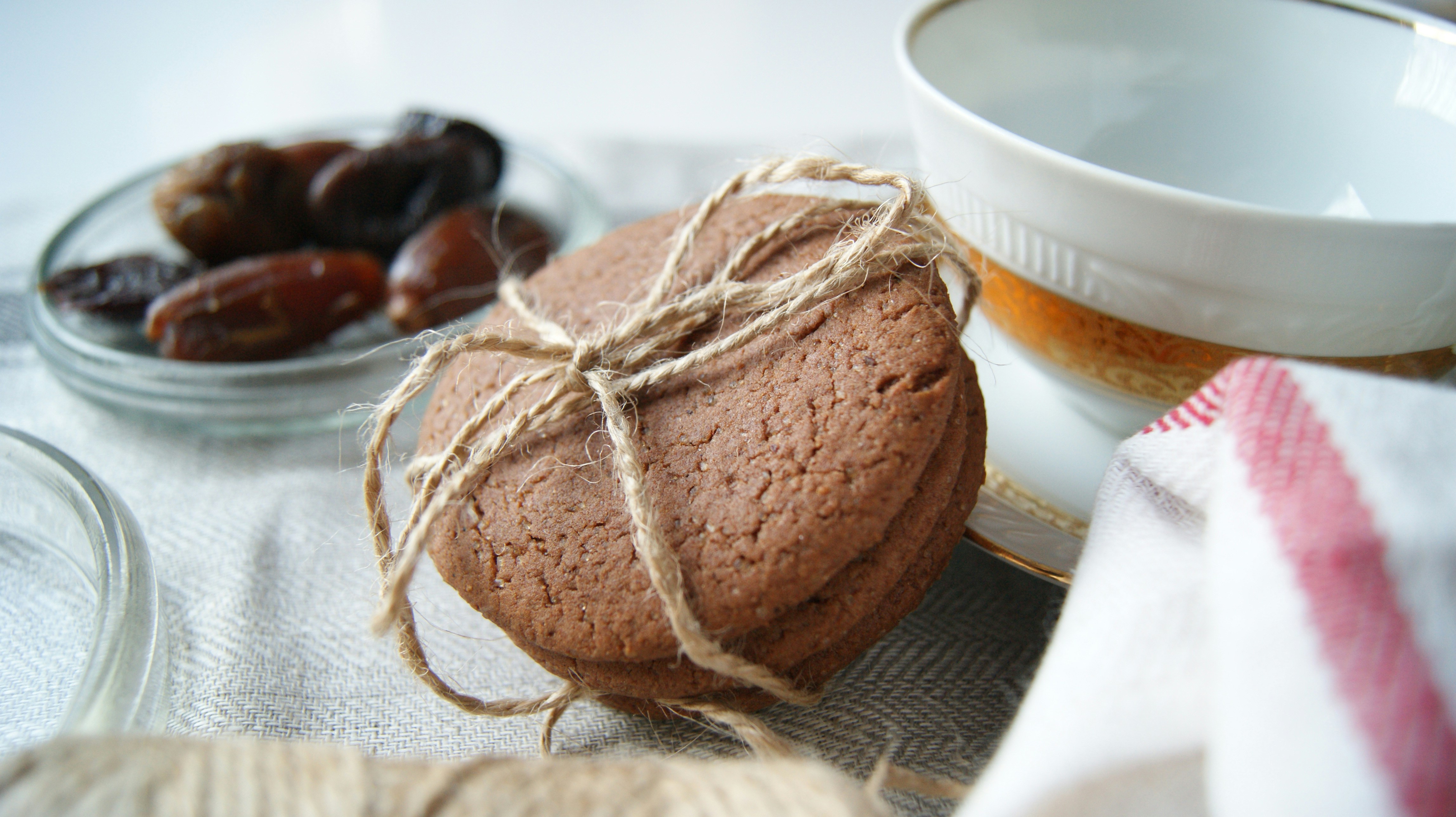Brown cookies tied with twine beside a white bowl and dates on a cloth.