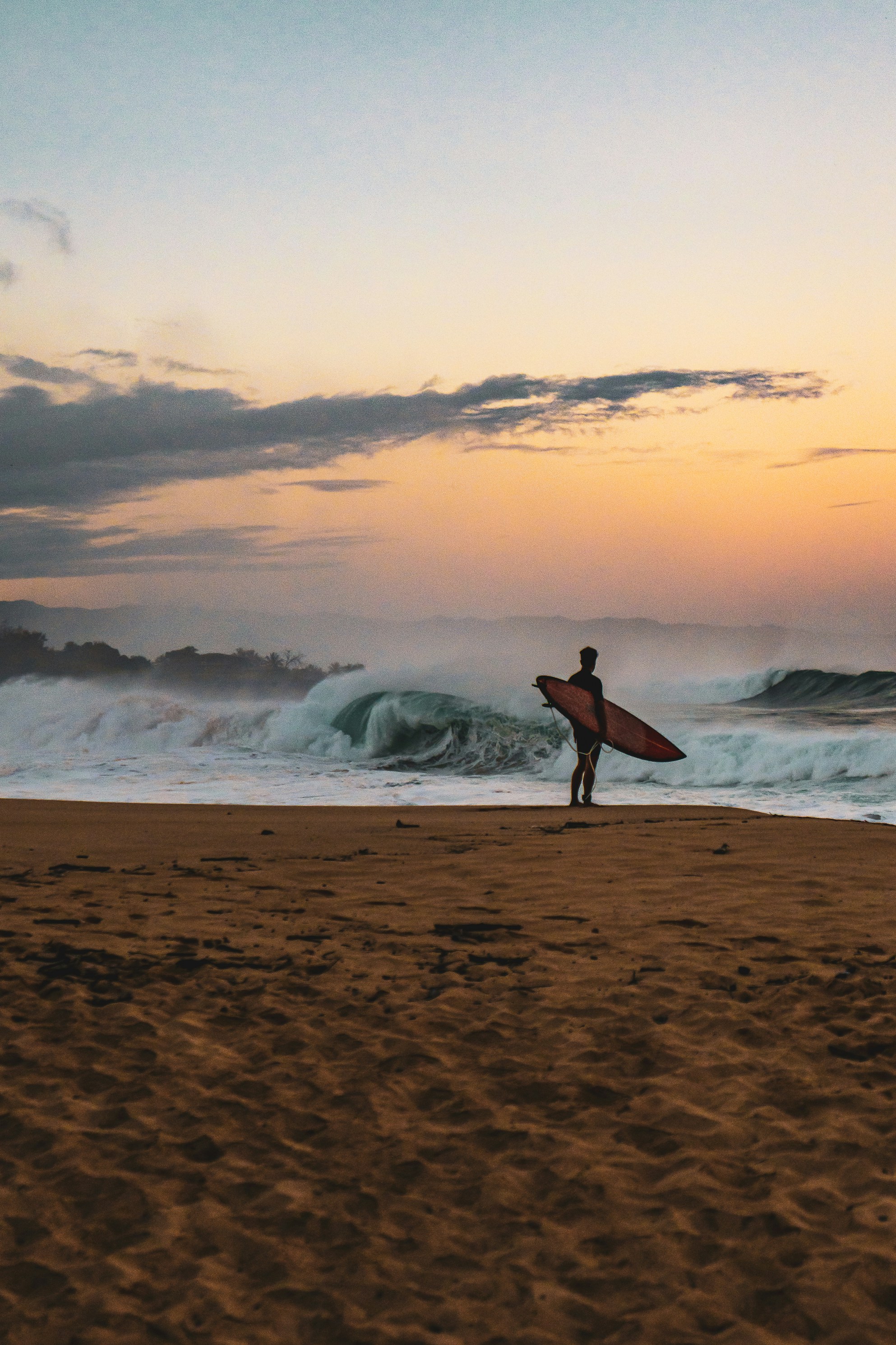 Silhouette of a surfer holding a surfboard on the beach at dawn, with crashing waves and a colorful sky in the background.