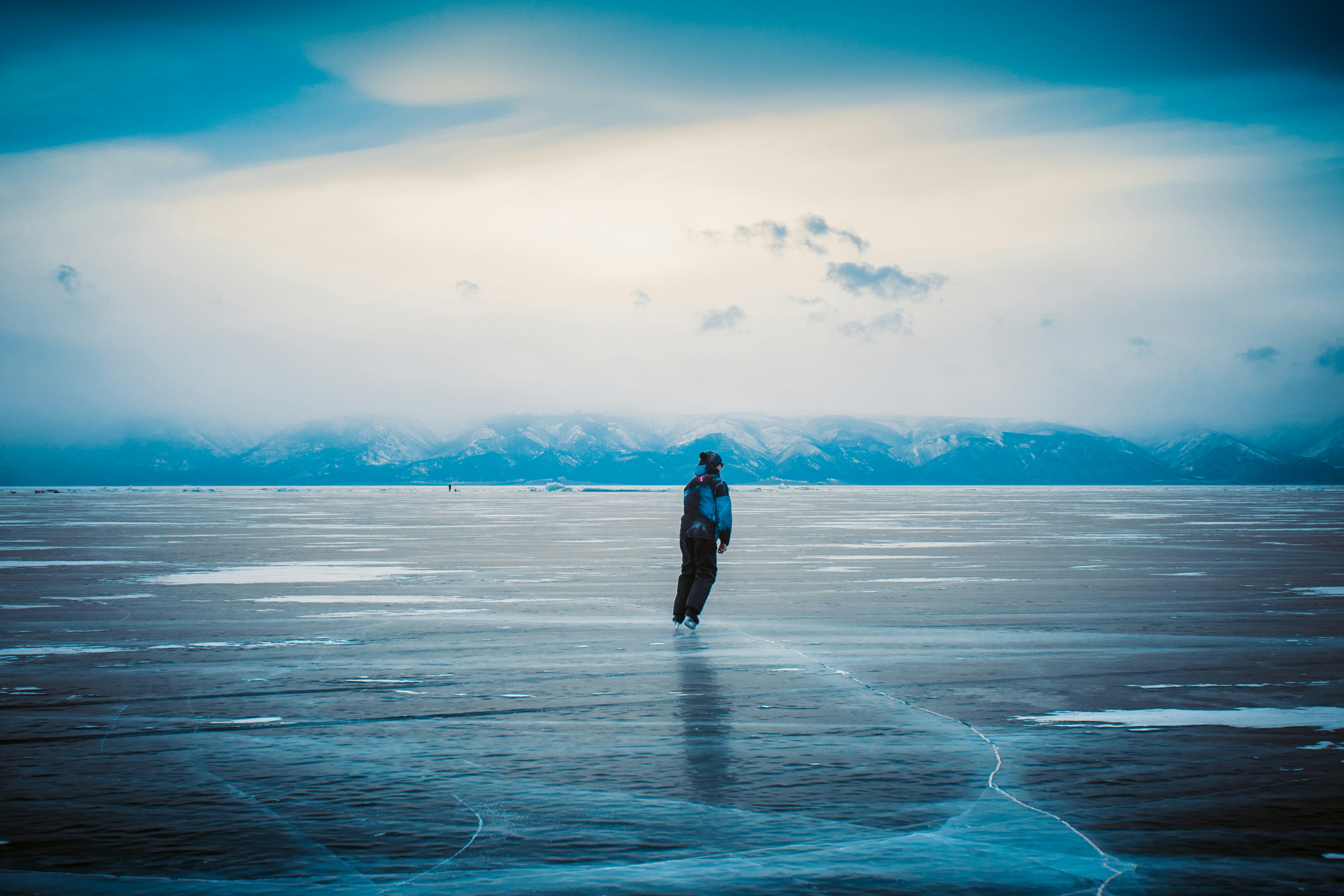 Person standing on a vast frozen lake under a cloudy sky with distant mountains.