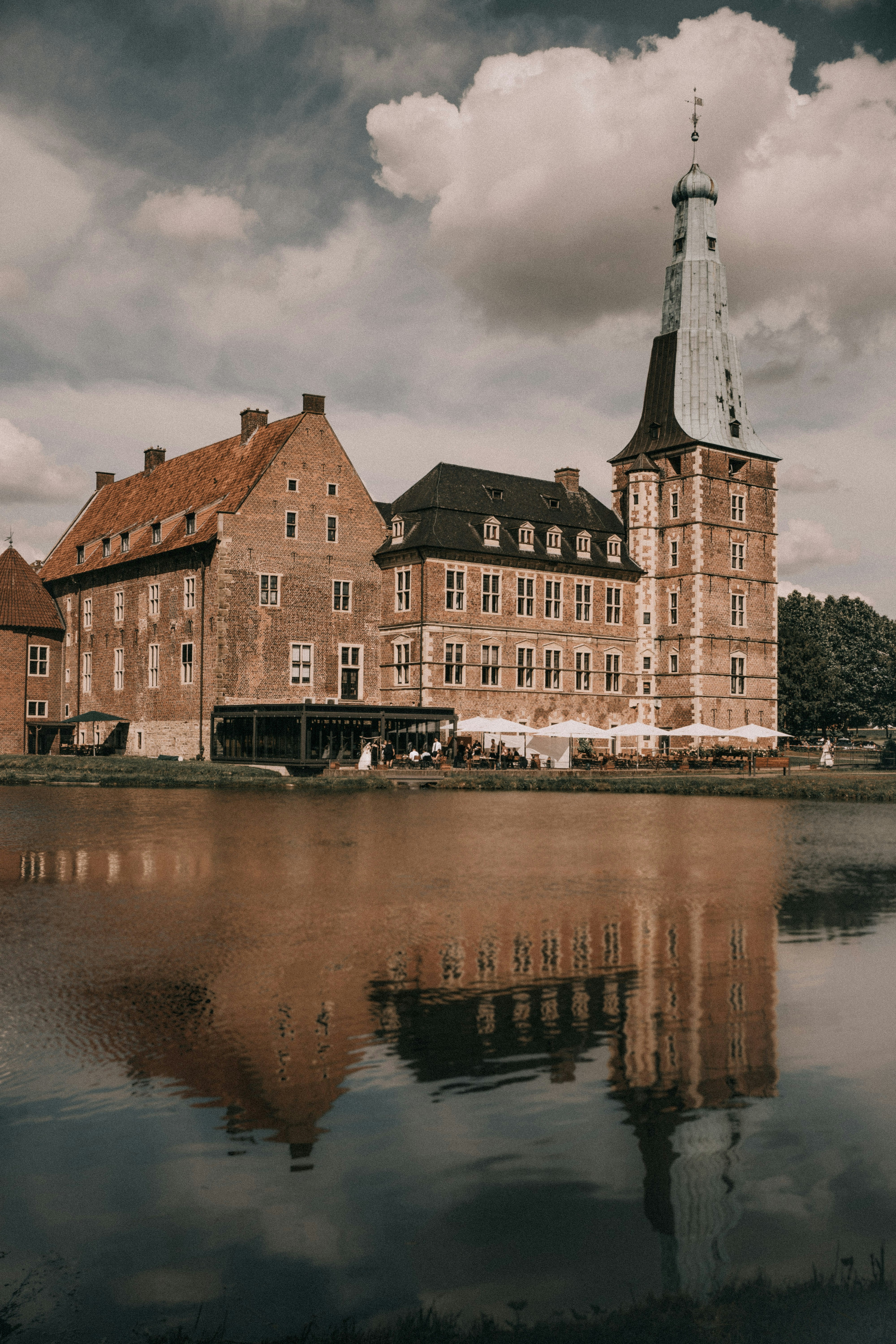 Historic brick building with a tall spire reflected in a tranquil lake under a cloudy sky.