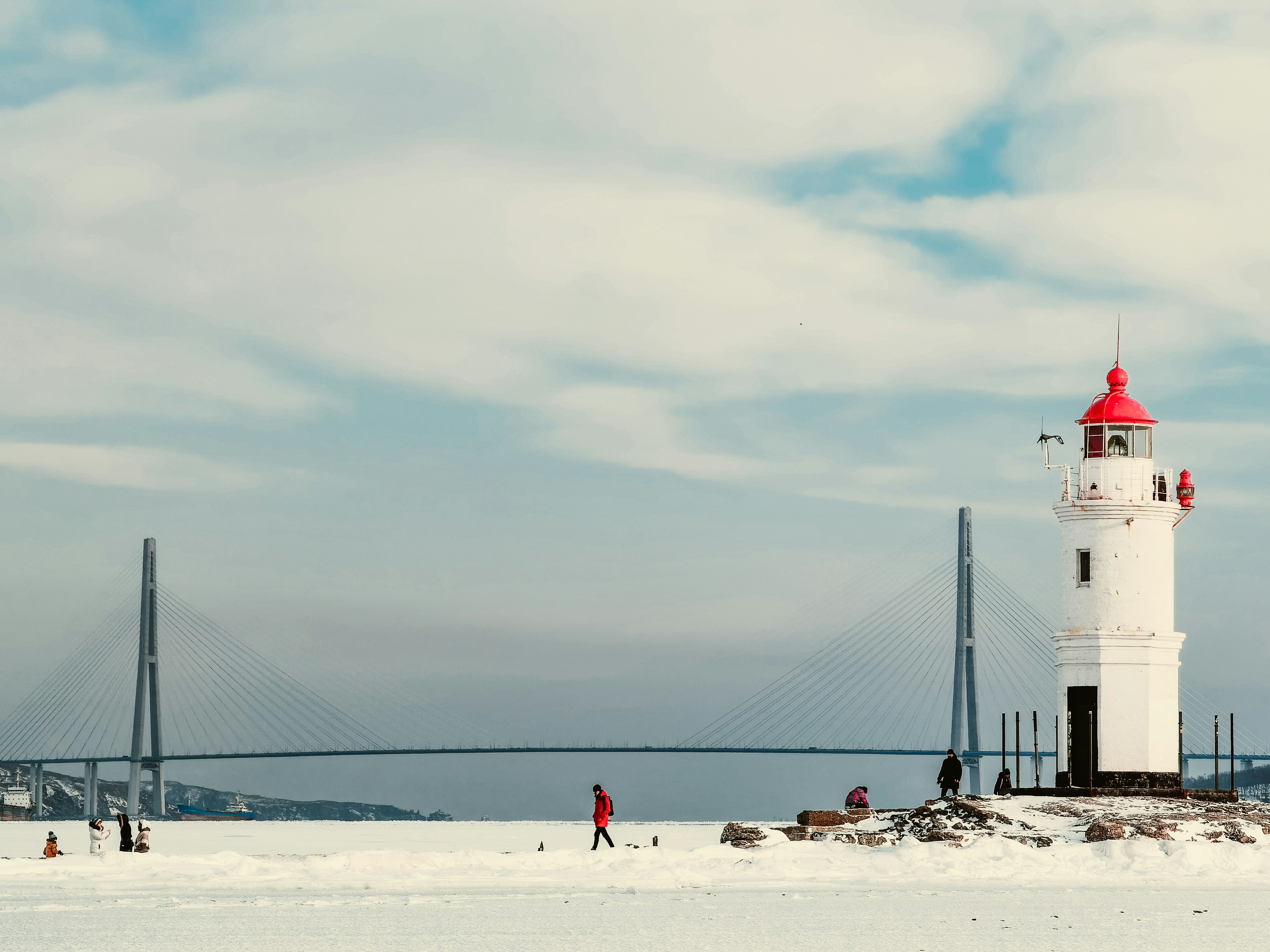 A solitary lighthouse stands against a backdrop of a vast, frozen landscape, with a modern bridge stretching across the distance. People traverse the snowy expanse, adding life to the serene scene.