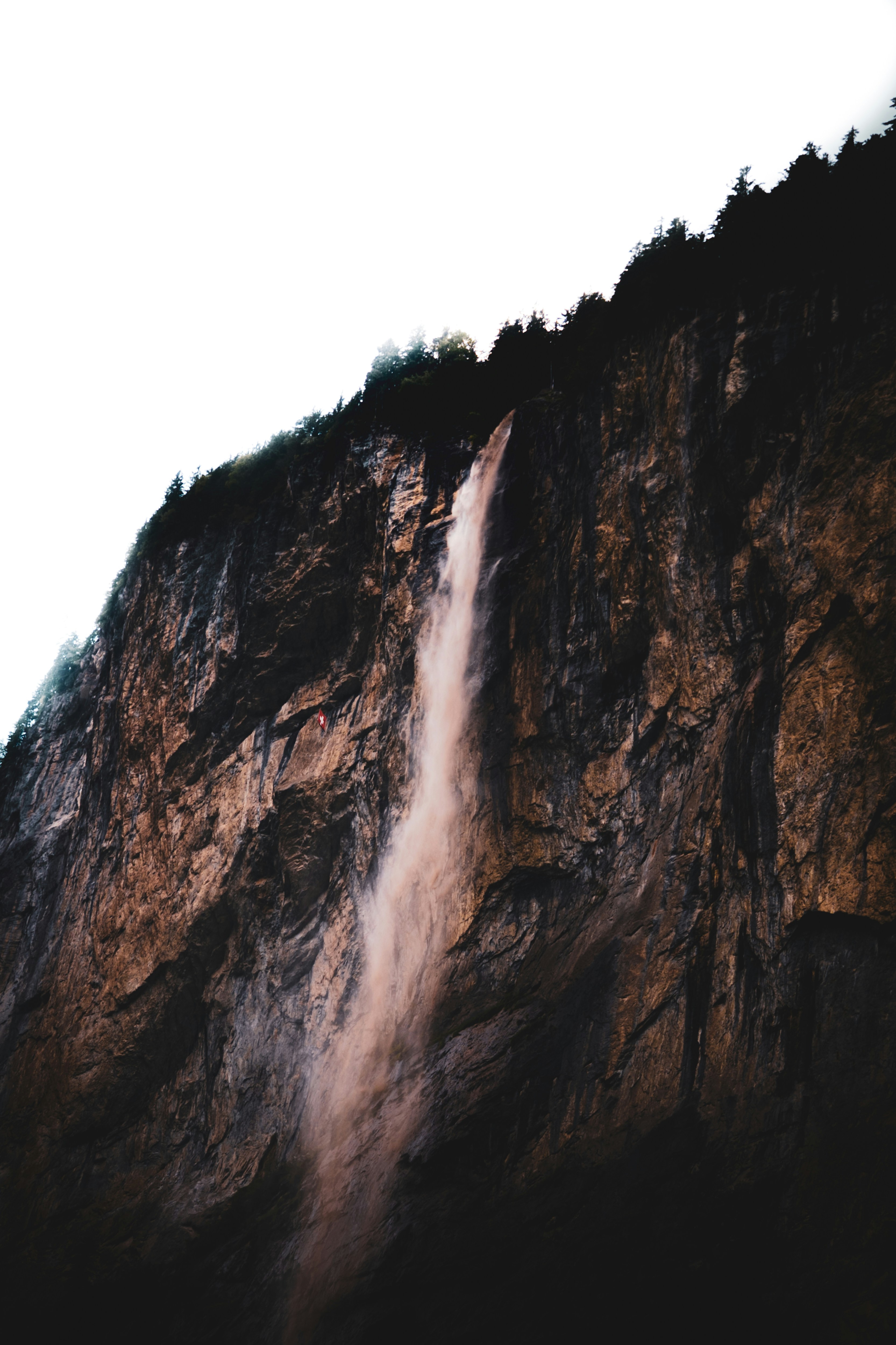 Waterfall cascading down a rugged cliffside, surrounded by dense forest. The interplay of light and shadow enhances the dramatic landscape.