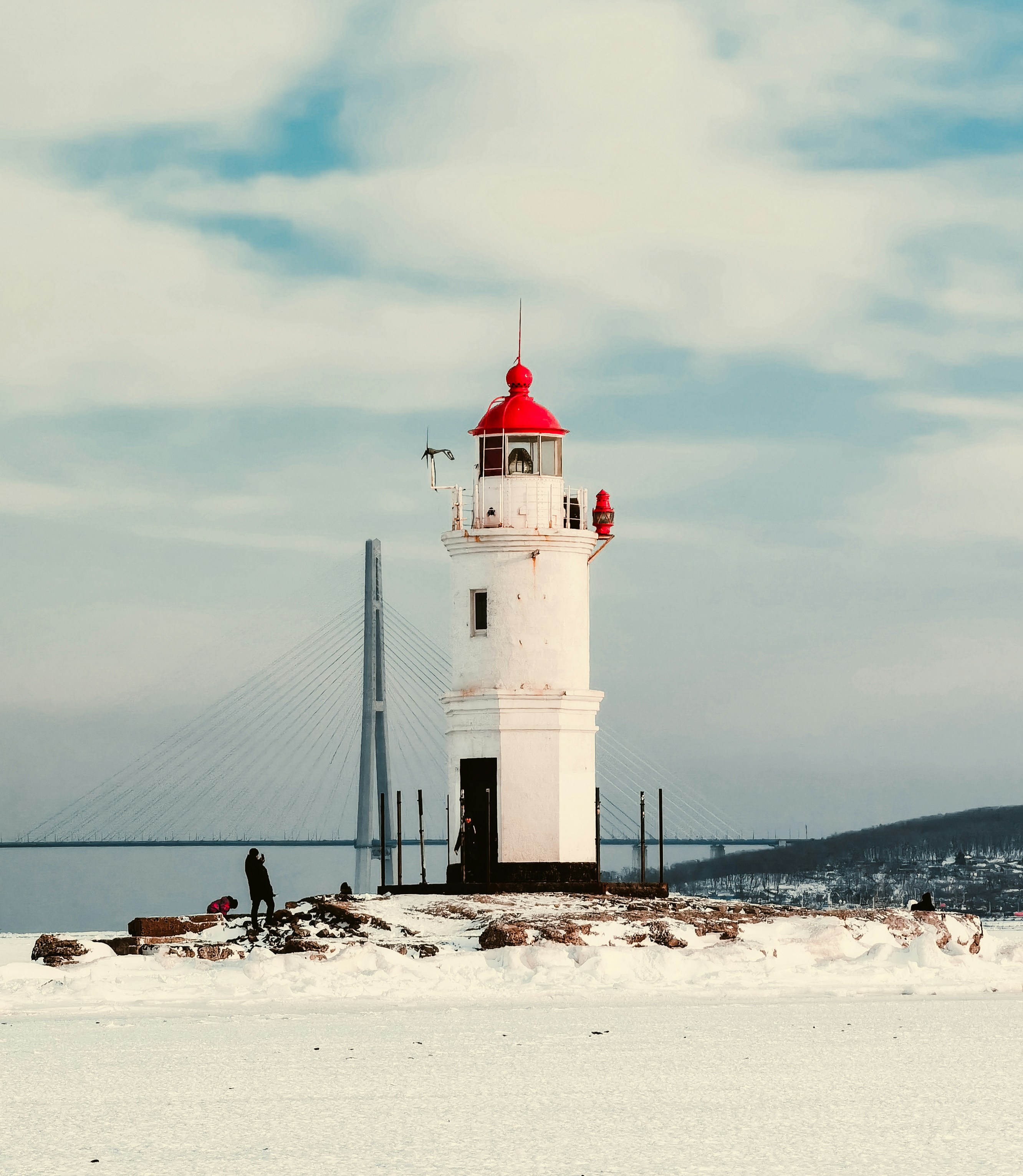 A solitary lighthouse stands on a snowy island, framed by a distant bridge under a soft winter sky.