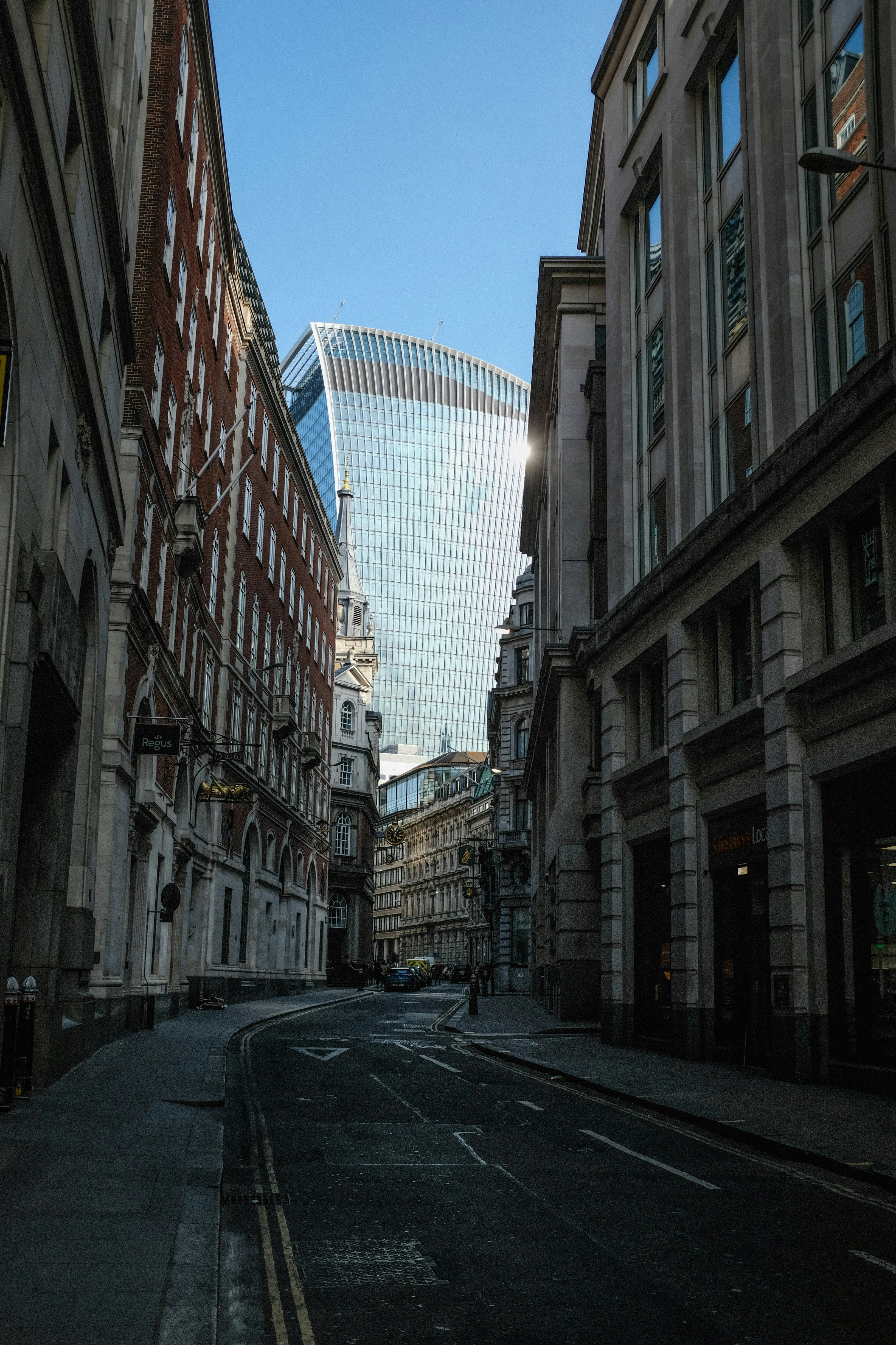 Narrow street framed by historical buildings leads to a modern skyscraper reflecting the sky. The scene highlights the juxtaposition of old and new architecture.