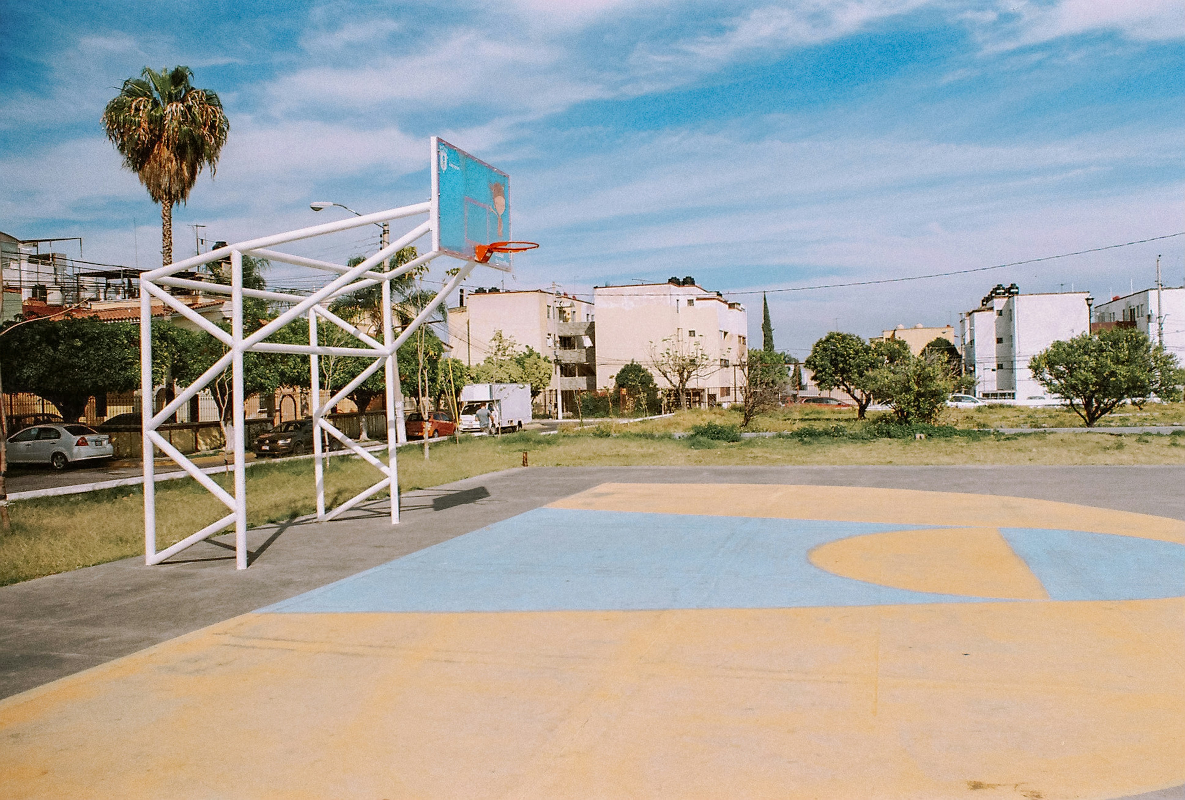Abandoned basketball court under a clear sky, featuring a lone hoop and vibrant painted surface. The surrounding greenery adds a touch of nature to the urban setting.