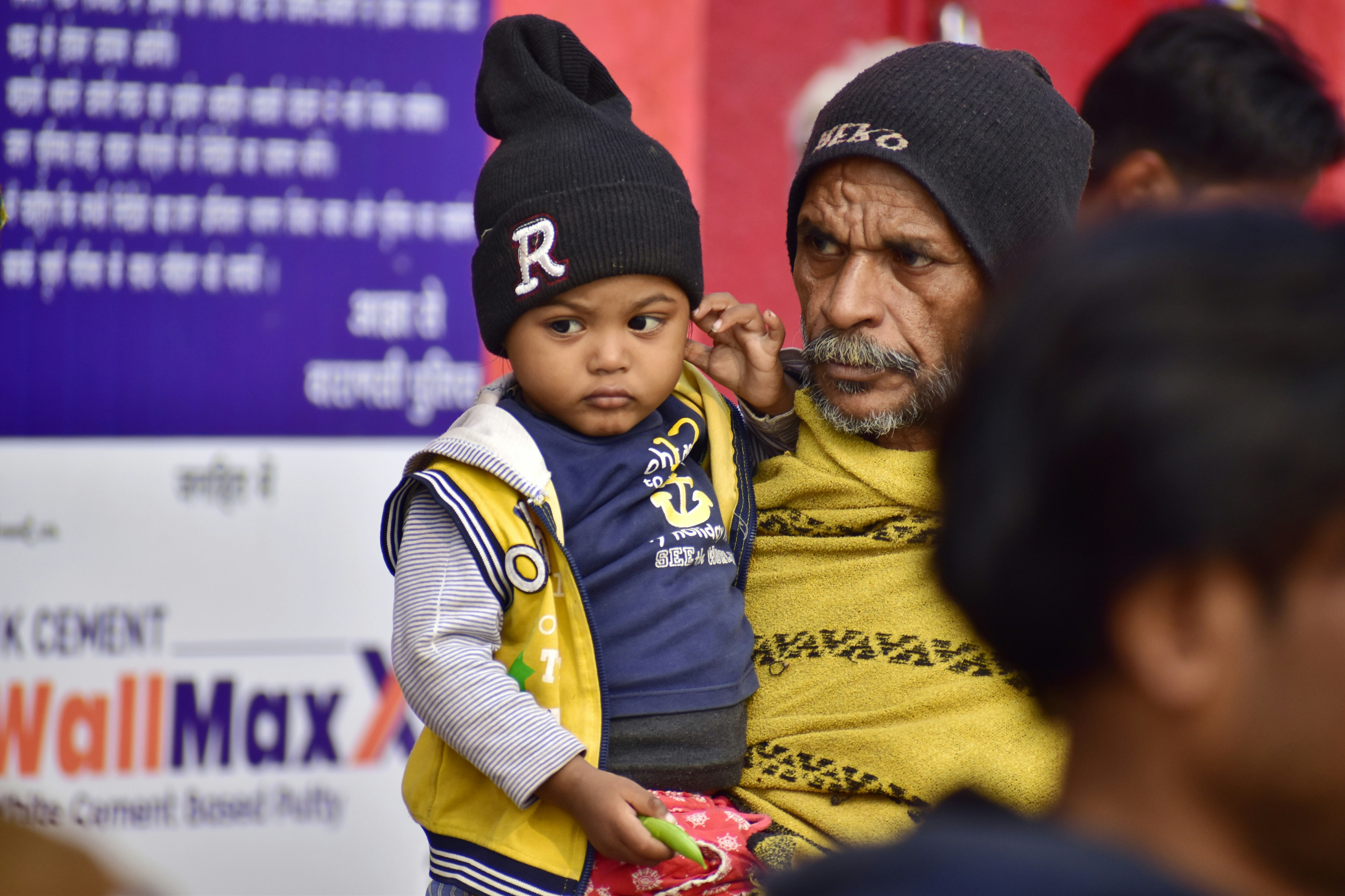 An elderly man holds a young child close, both dressed warmly, amidst a vibrant backdrop of colorful signage.