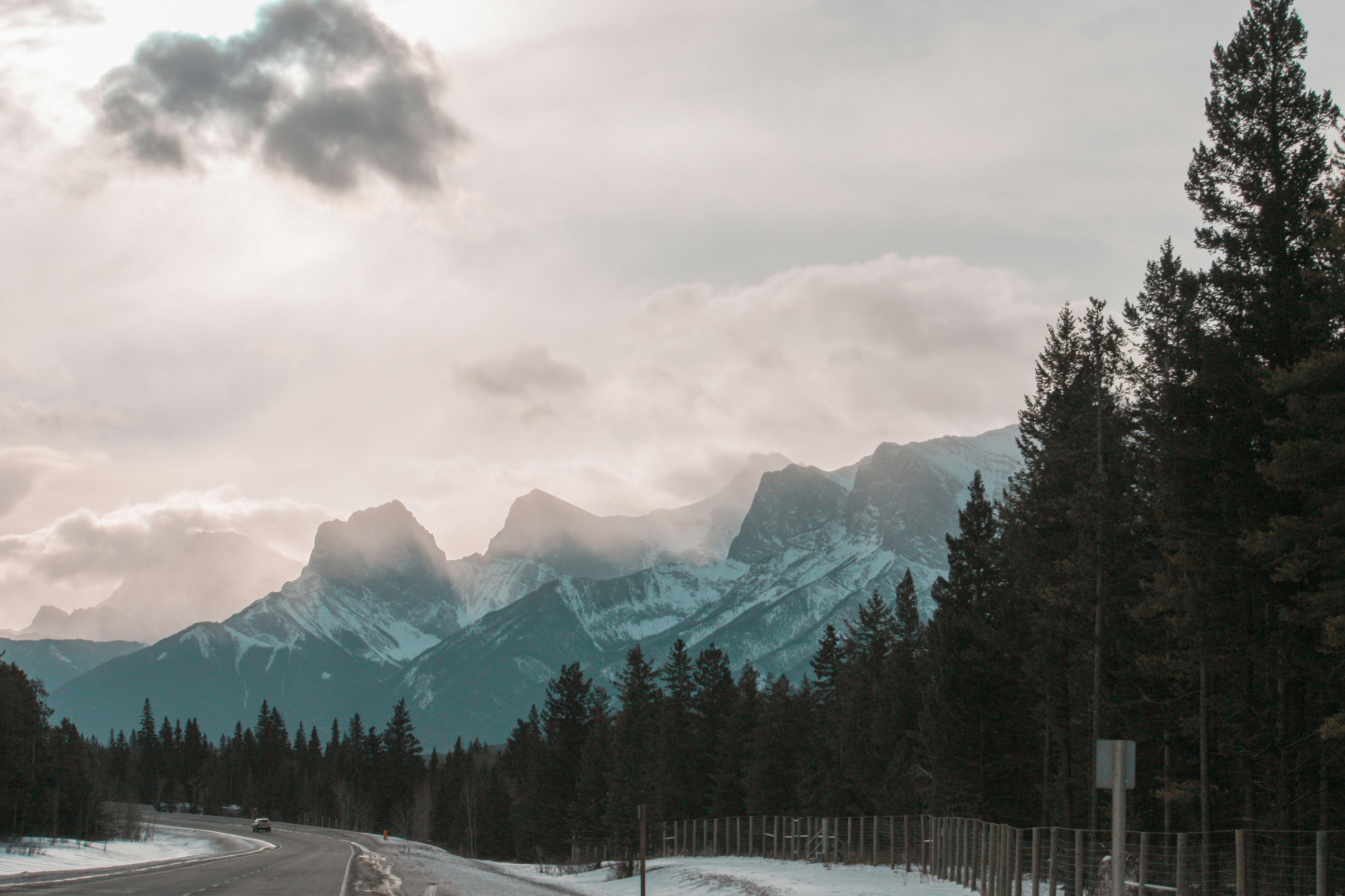 Snow-dusted mountains under a cloudy sky with a winding road flanked by towering evergreens.