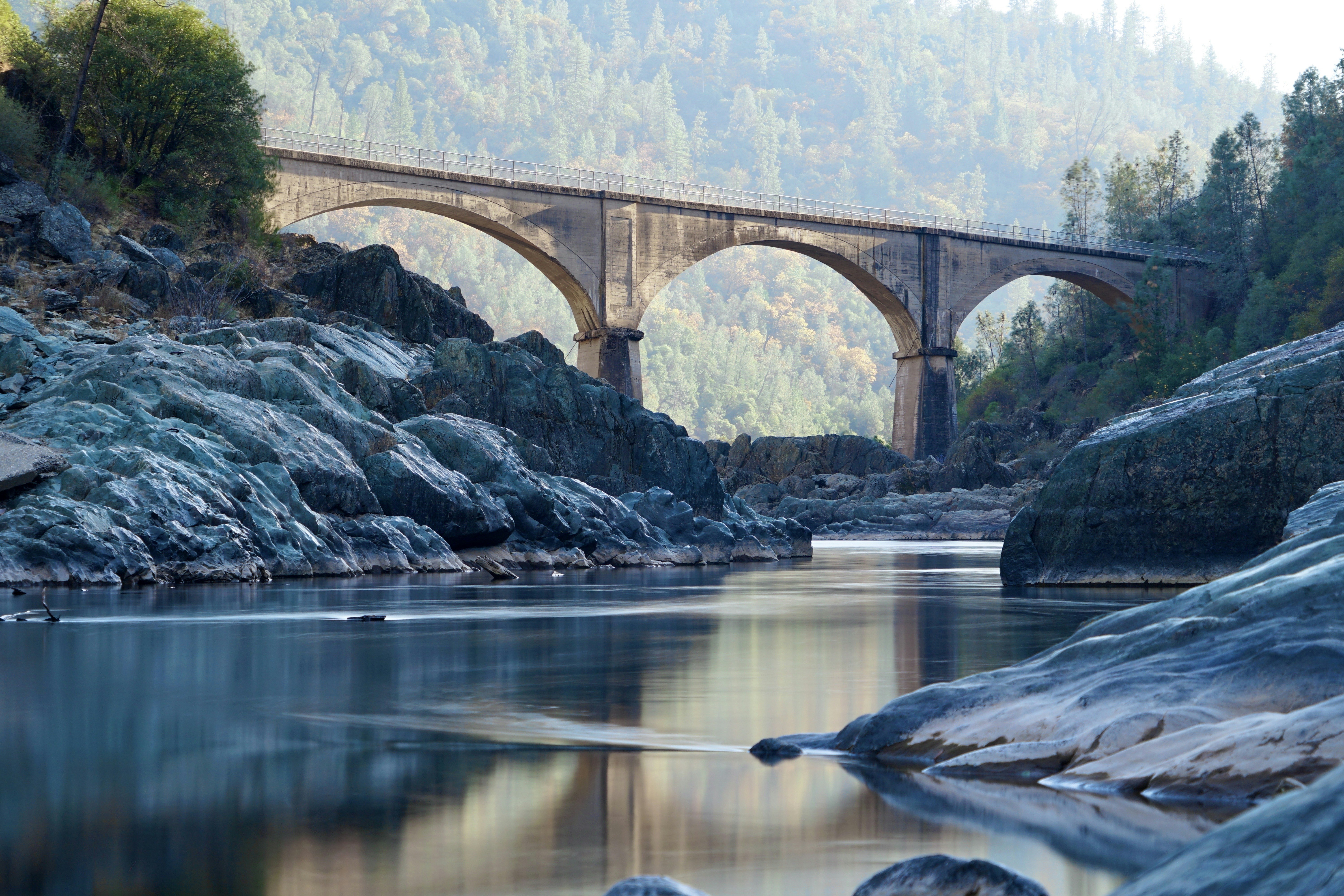 Arch bridge spans a tranquil river flanked by rocky banks and lush hillsides.