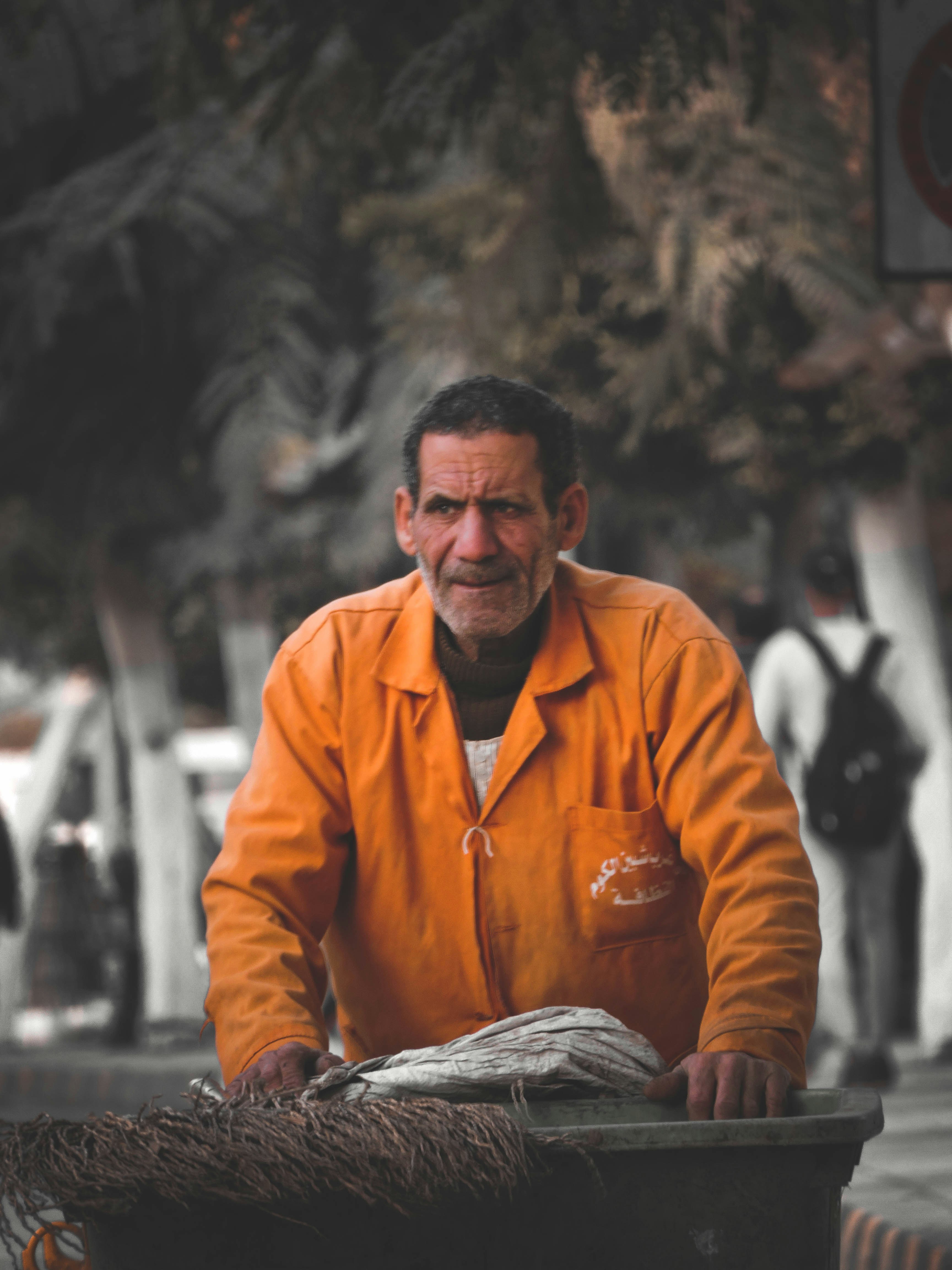 A man in an orange work jacket leans over a vendor cart on a city street, his hands resting on the edge. Pedestrians and foliage blur in the background, keeping the focus on the subject.