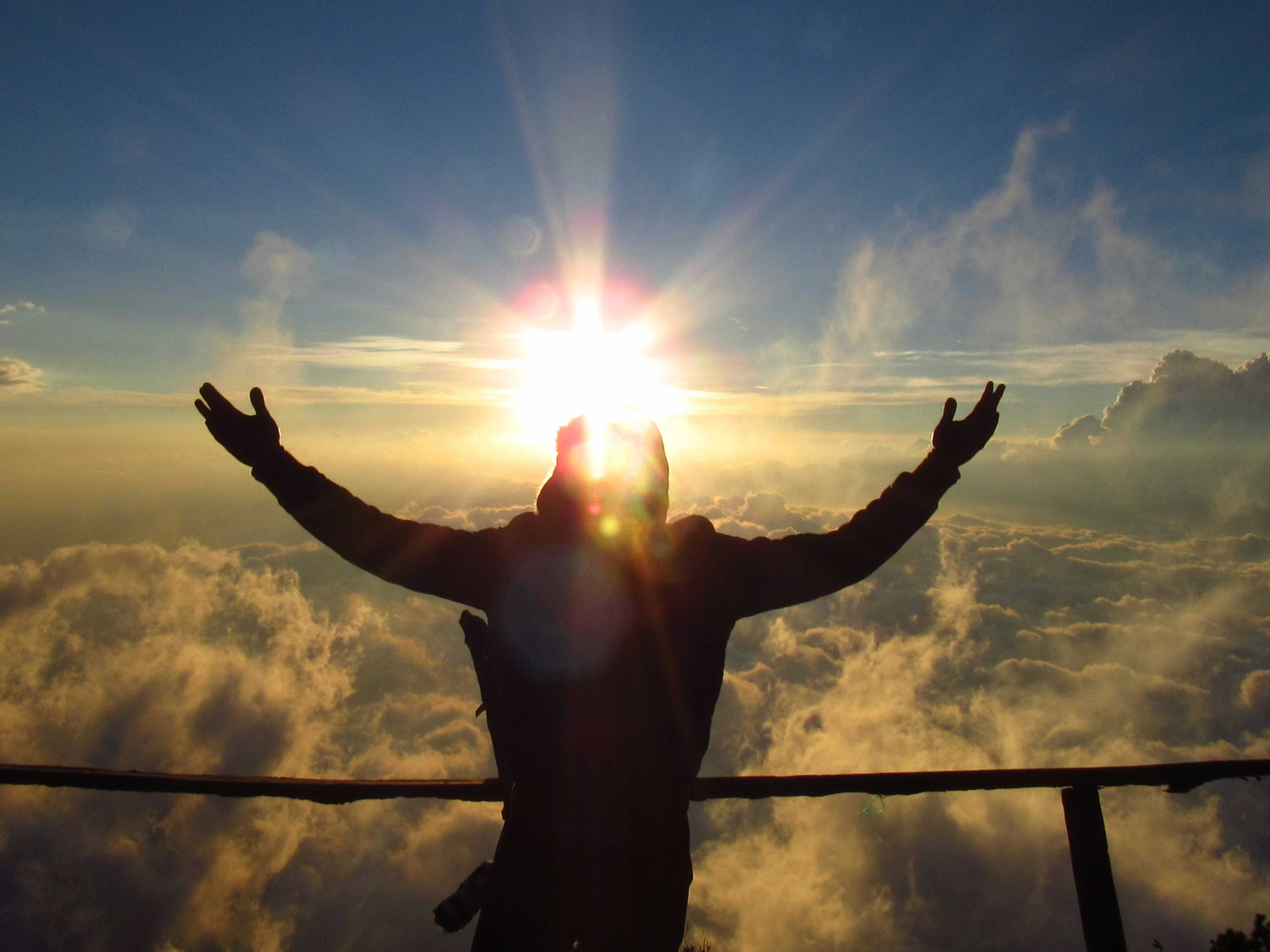 Silhouetted figure stands triumphantly on a ledge, arms outstretched towards the sun rising above a sea of clouds.