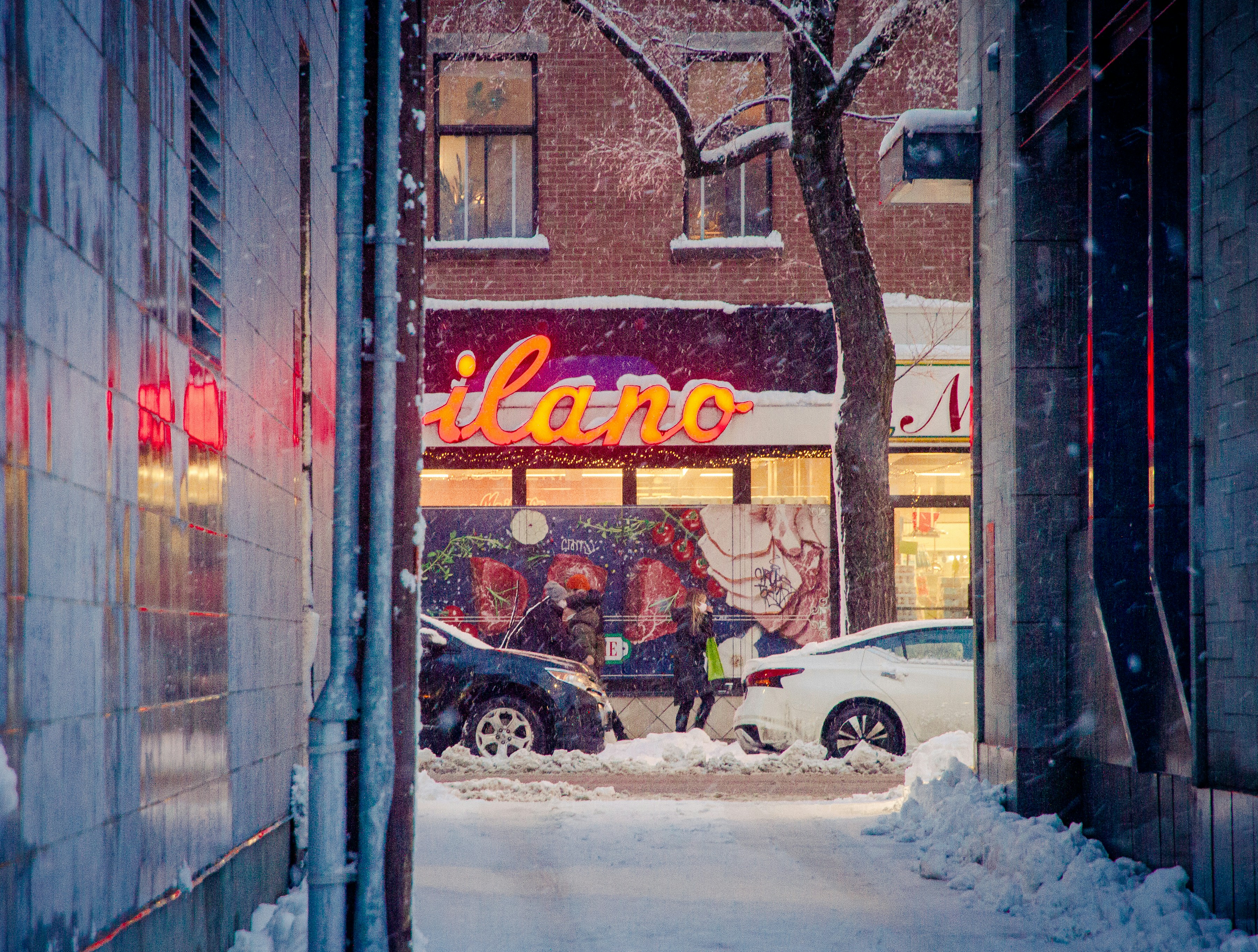 Snow-dusted alley frames a vibrant storefront with neon sign during snowfall.