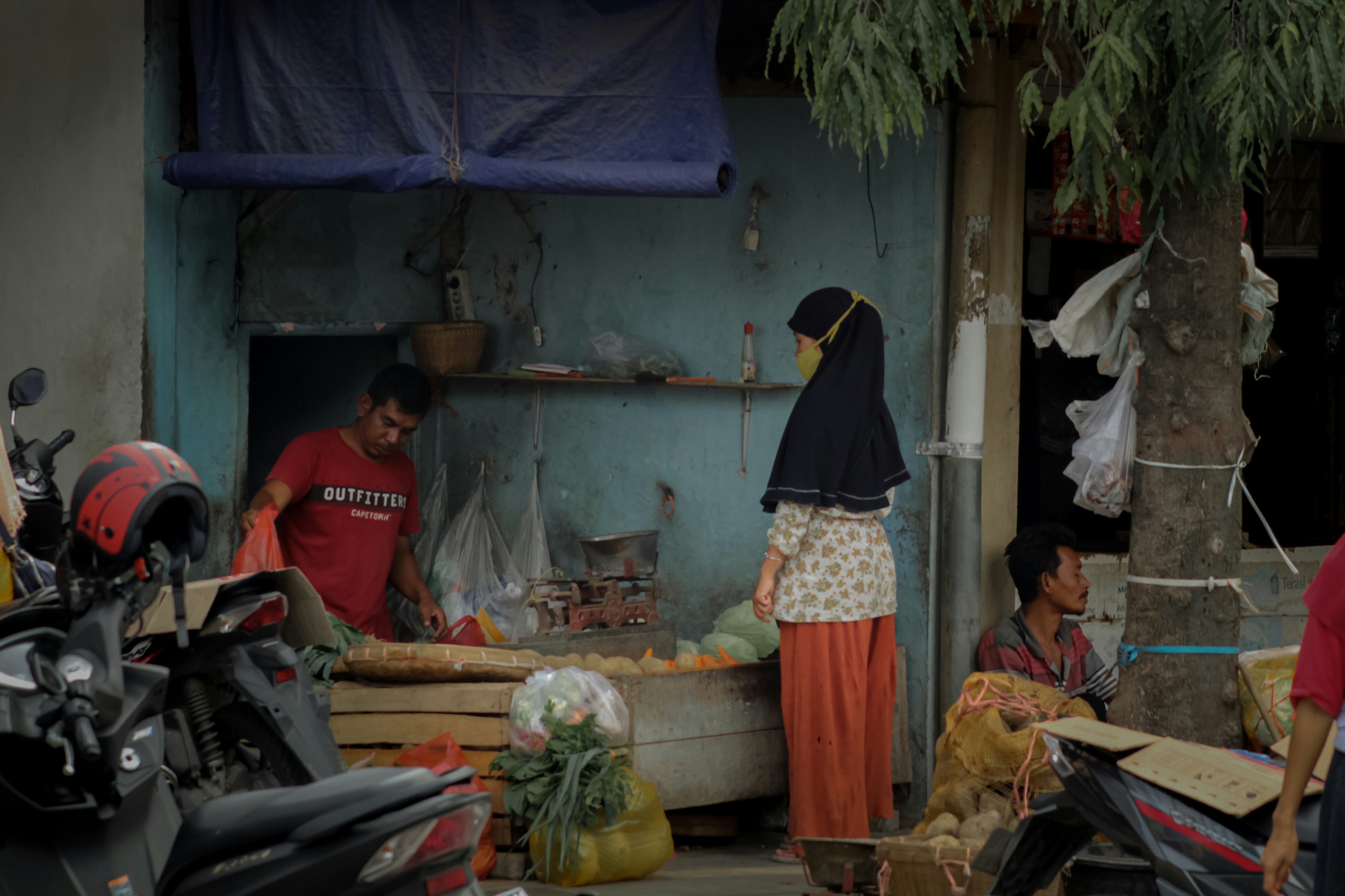 Vendors and customers interact in a small market stall surrounded by motorbikes and goods.