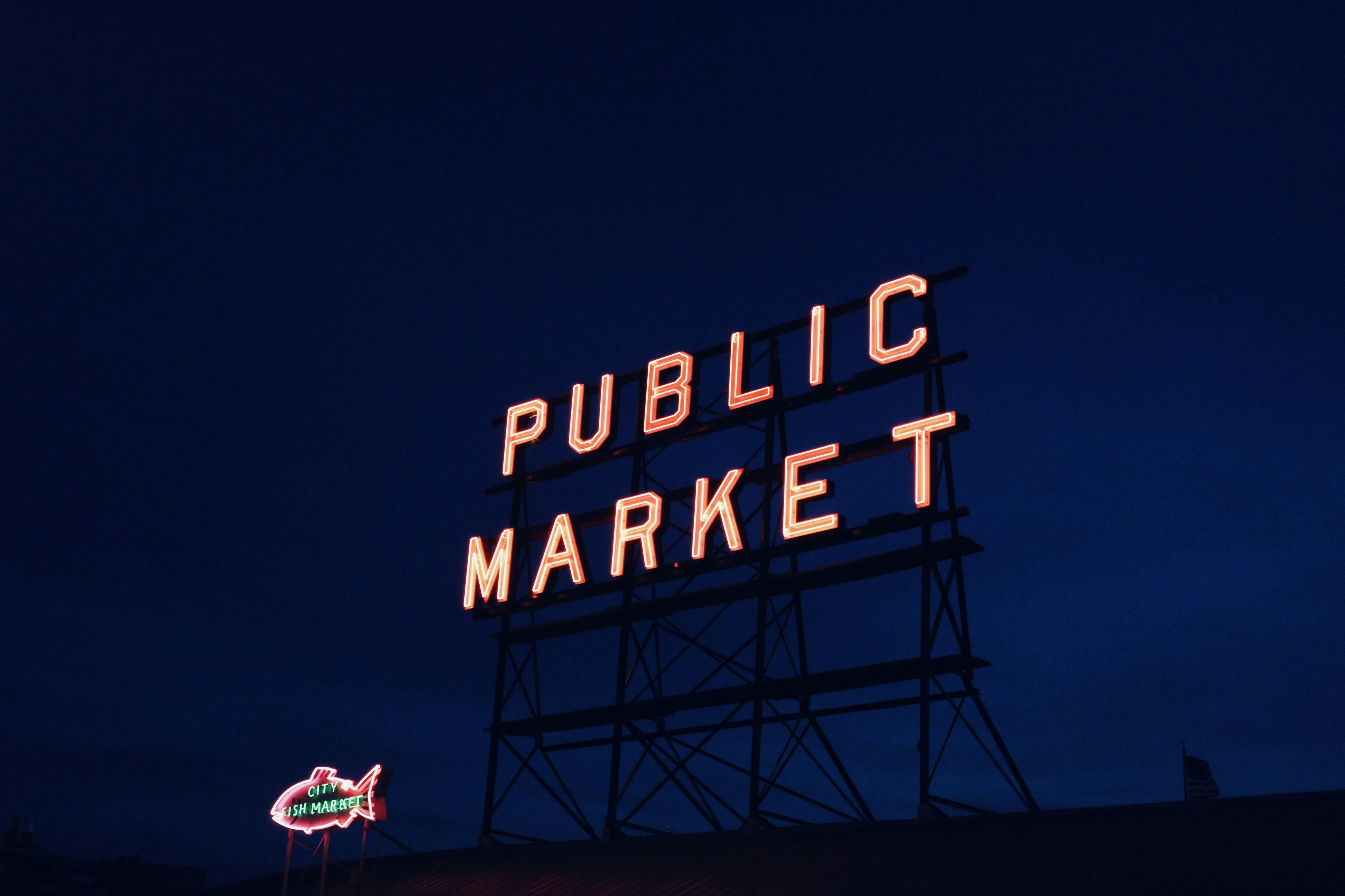 Illuminated 'PUBLIC MARKET' sign glowing against a twilight sky, accompanied by a smaller fish logo. 