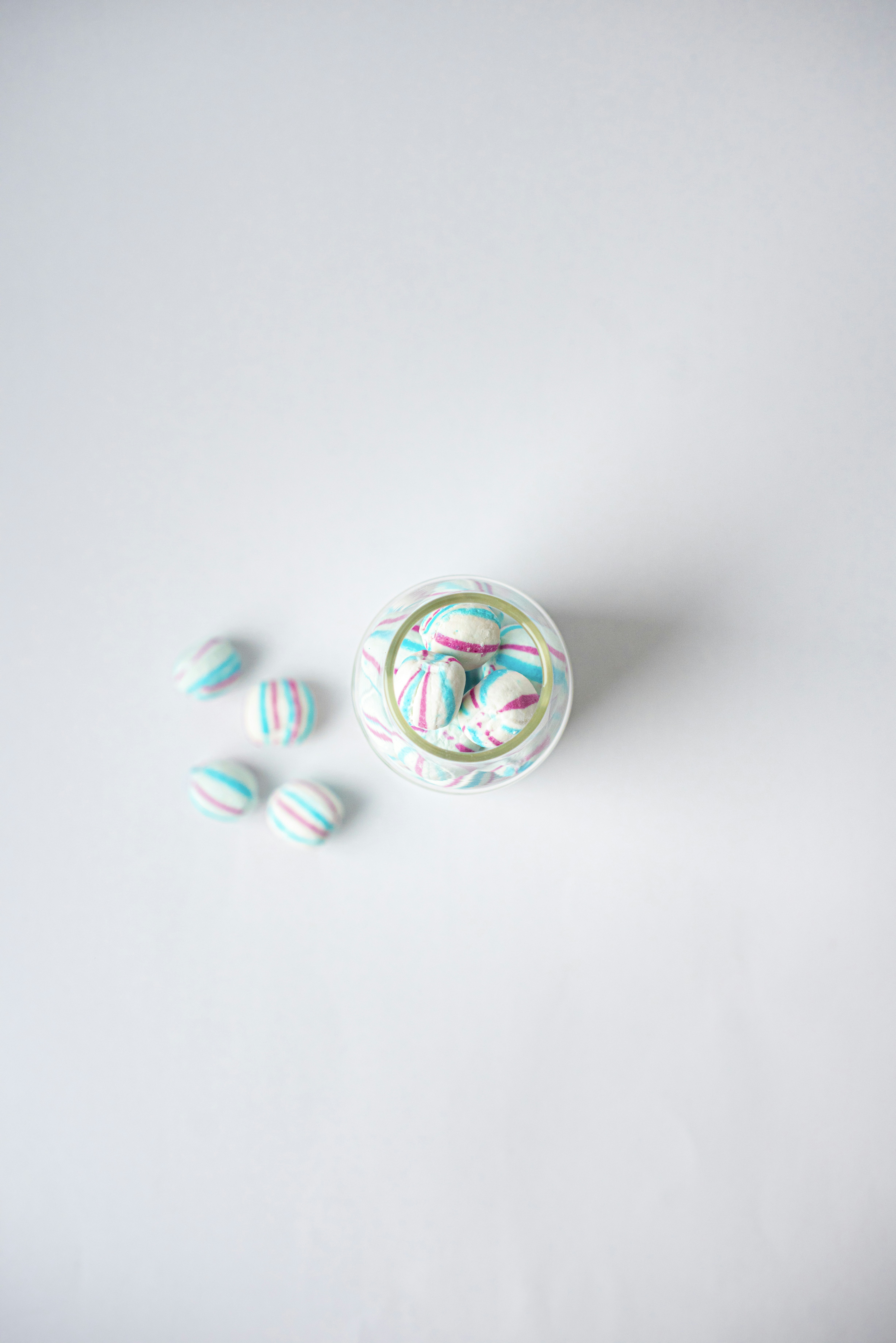 Colorful striped candies arranged in a glass jar, with a few scattered nearby on a soft white background.