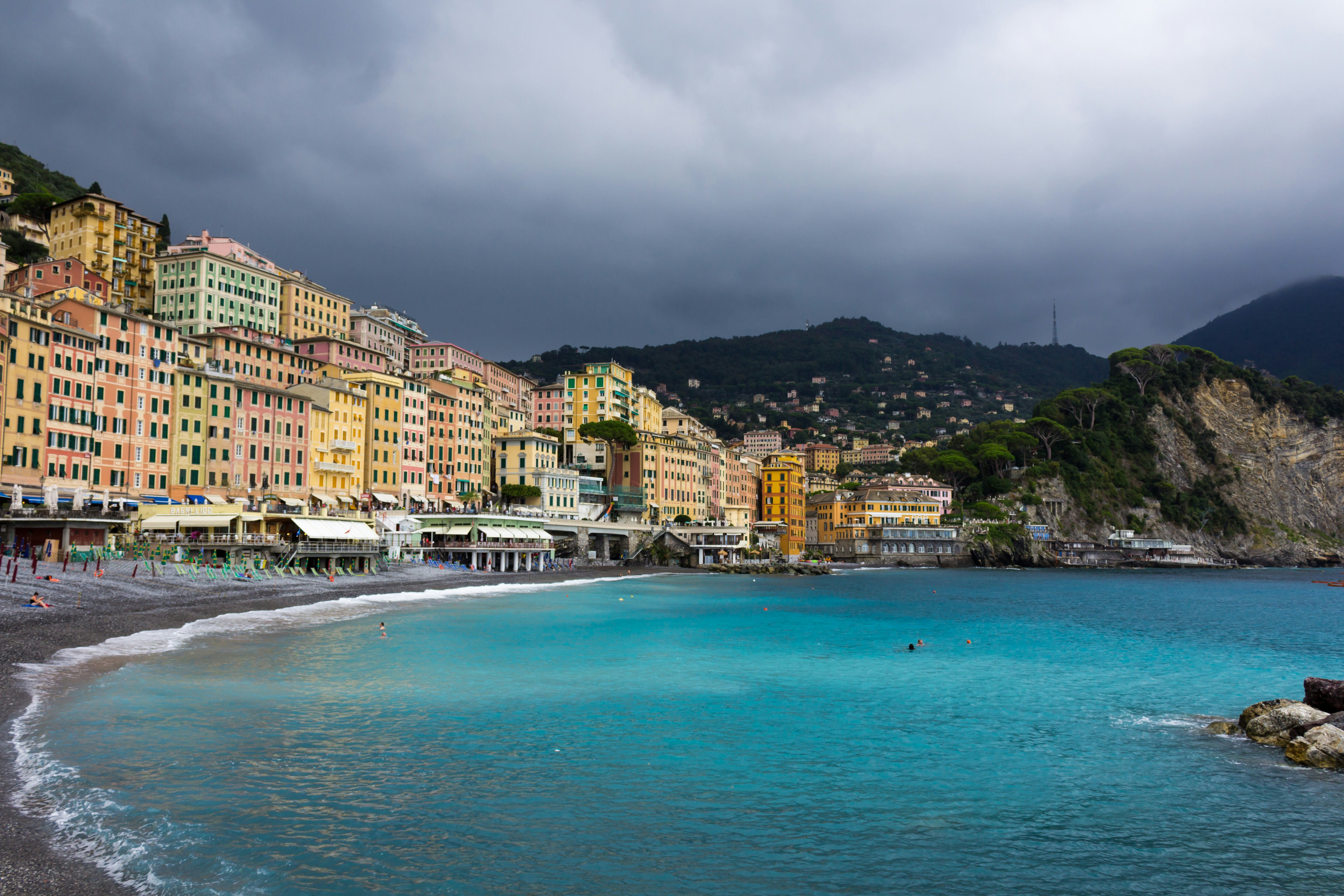 Vibrant coastal town with pastel-colored buildings lining the shore, under a dramatic sky. The turquoise water contrasts beautifully with the rocky landscape.