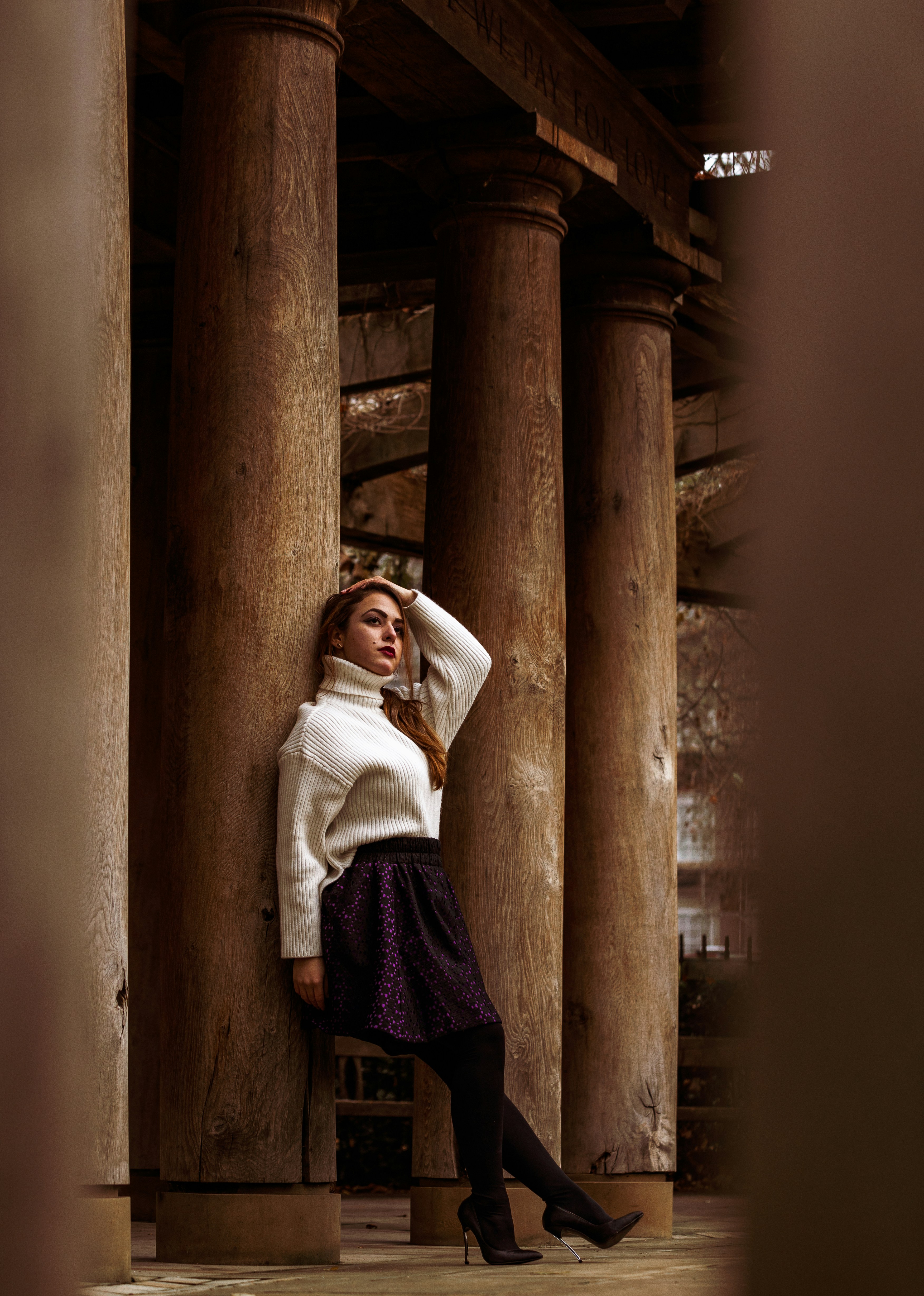 A woman in a white turtleneck and purple skirt leans against wooden columns, exuding a sense of calm and style in a serene setting.