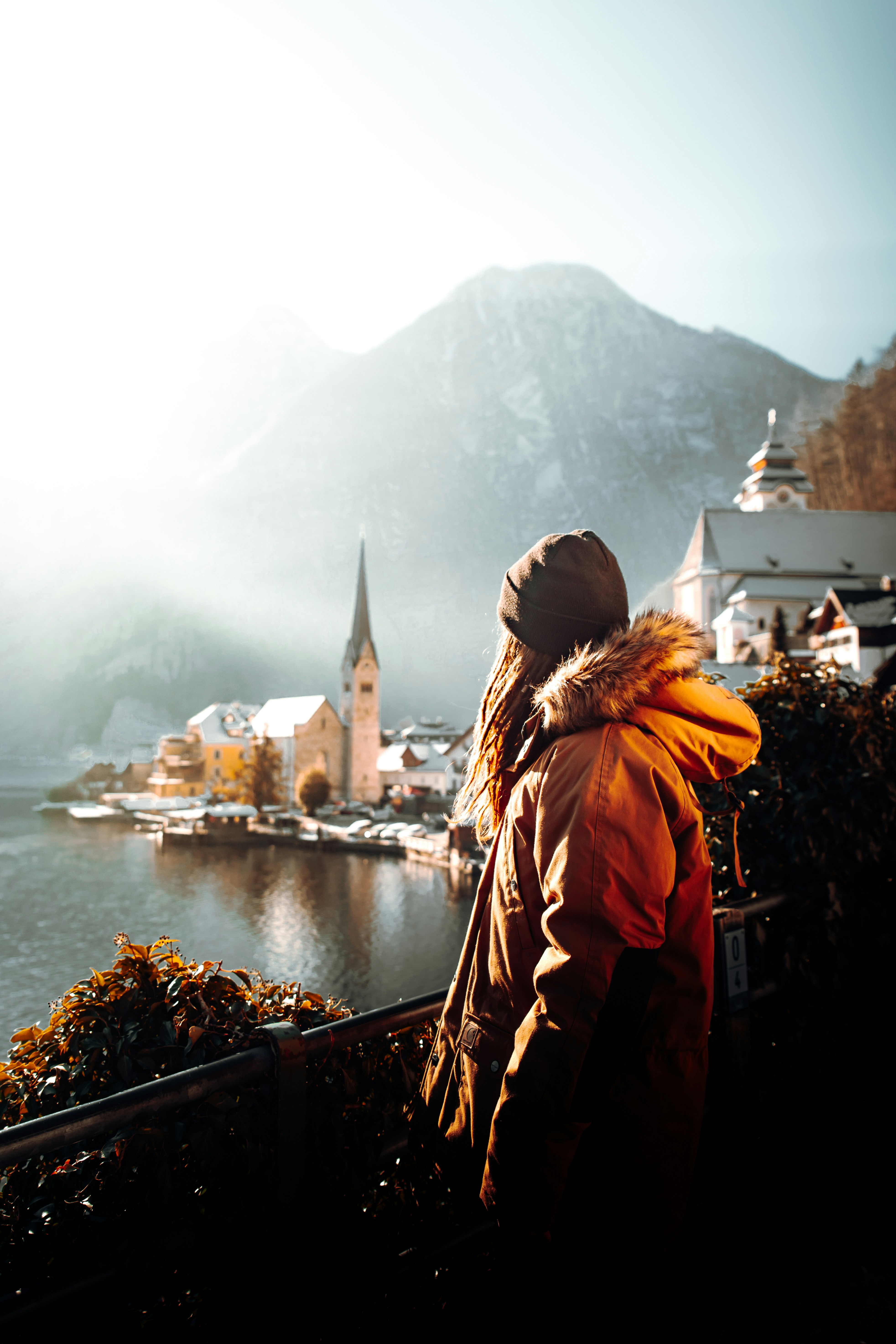A figure in an orange coat stands by a railing, gazing at a tranquil lakeside village surrounded by snow-capped mountains. The soft light creates a dreamy atmosphere.