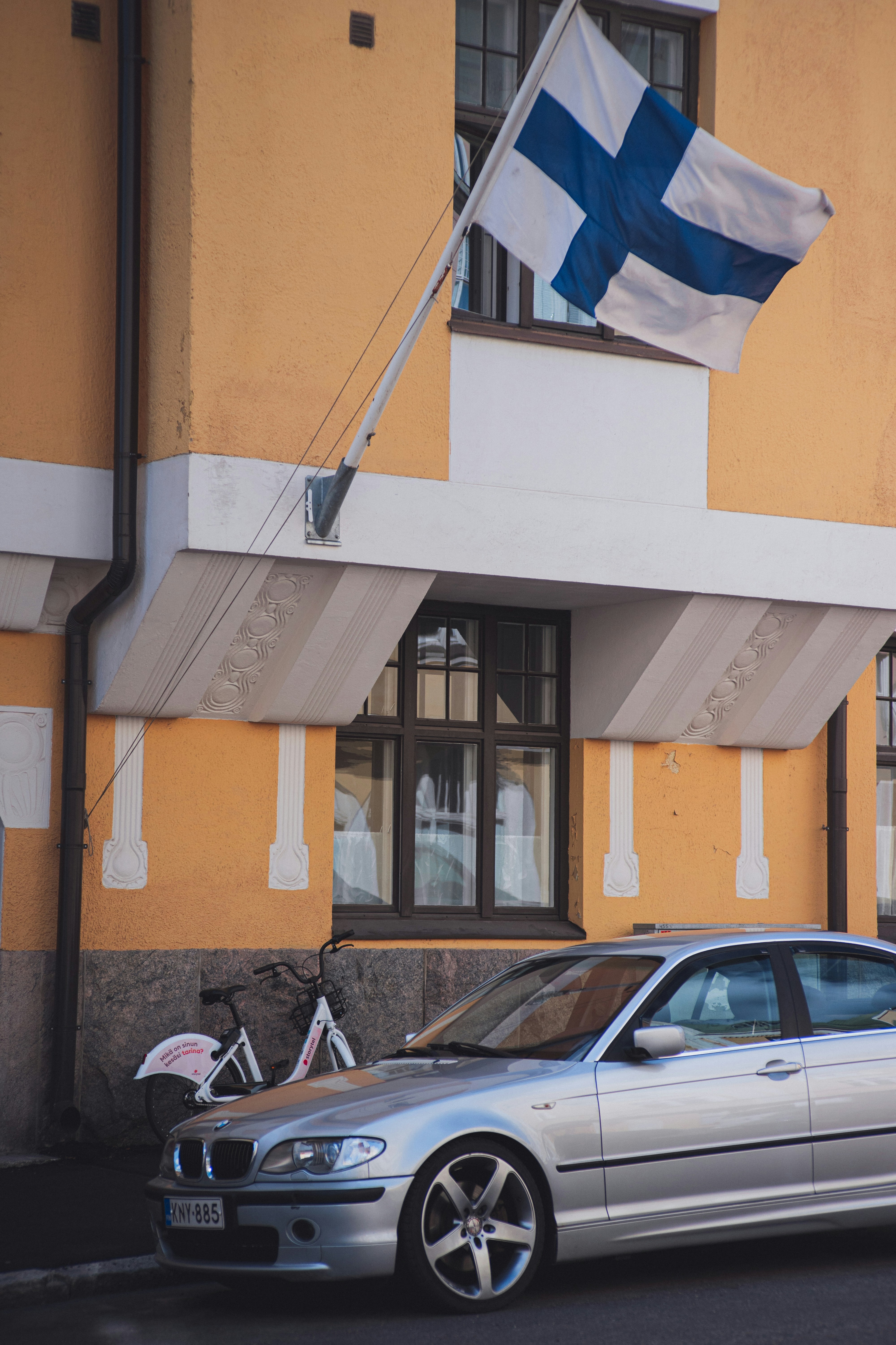 Nordic Cross in the City: Finnish Flag Draped Over a Sunlit Facade with a Silver BMW