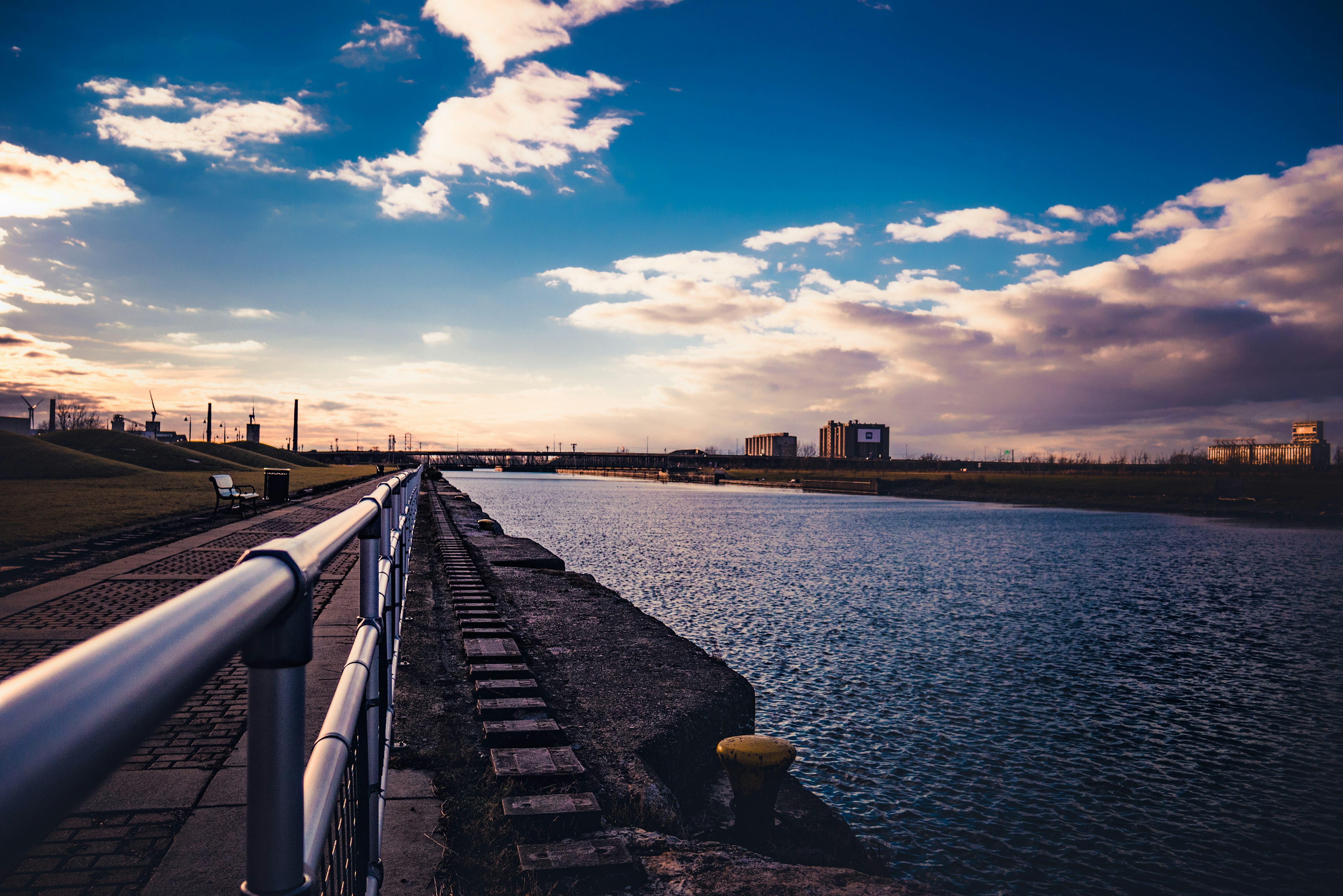 Riverside pathway with railing alongside calm water under a partly cloudy sky at sunset.