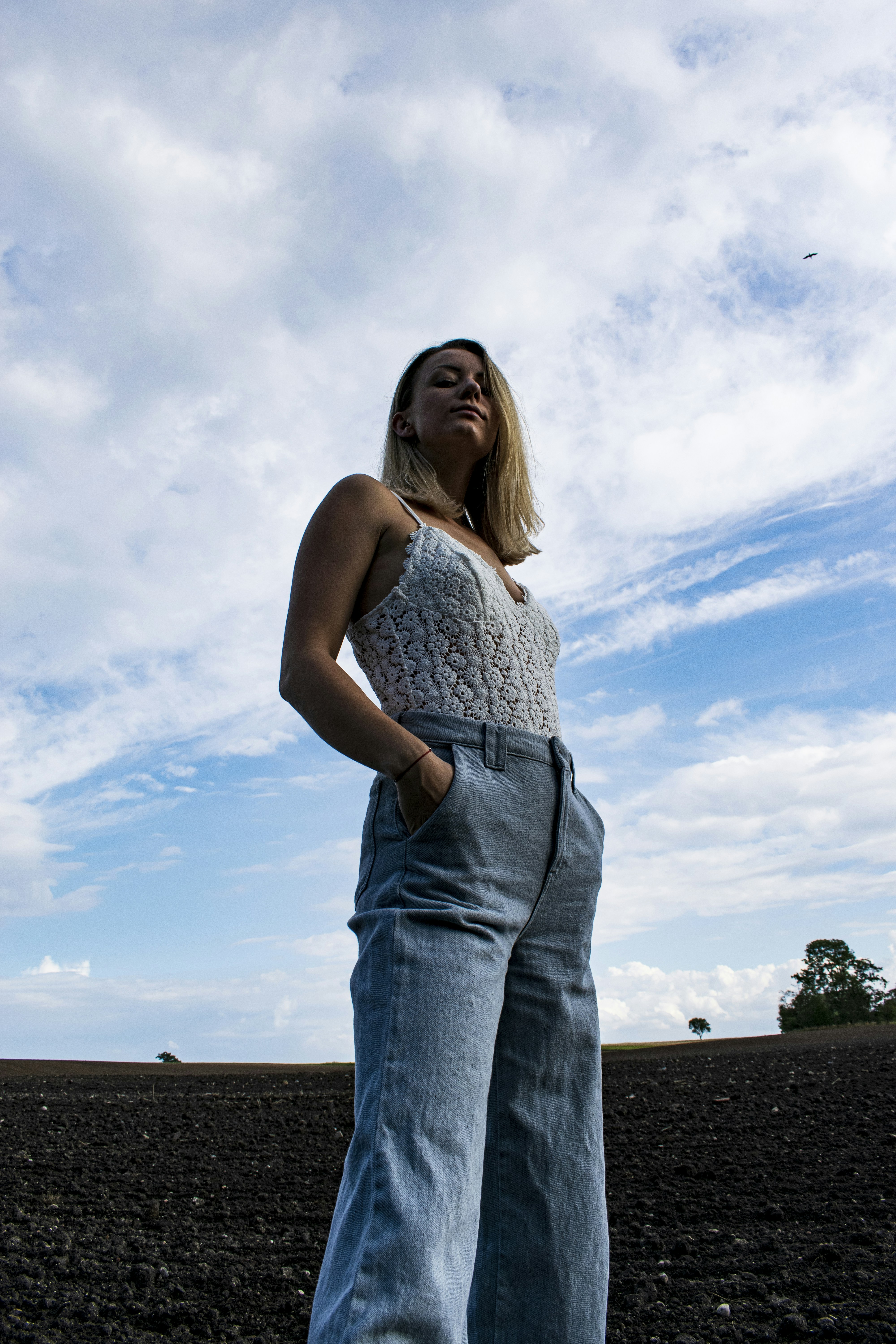 Woman in a textured top and wide-leg jeans stands confidently on a barren landscape under a cloudy sky.