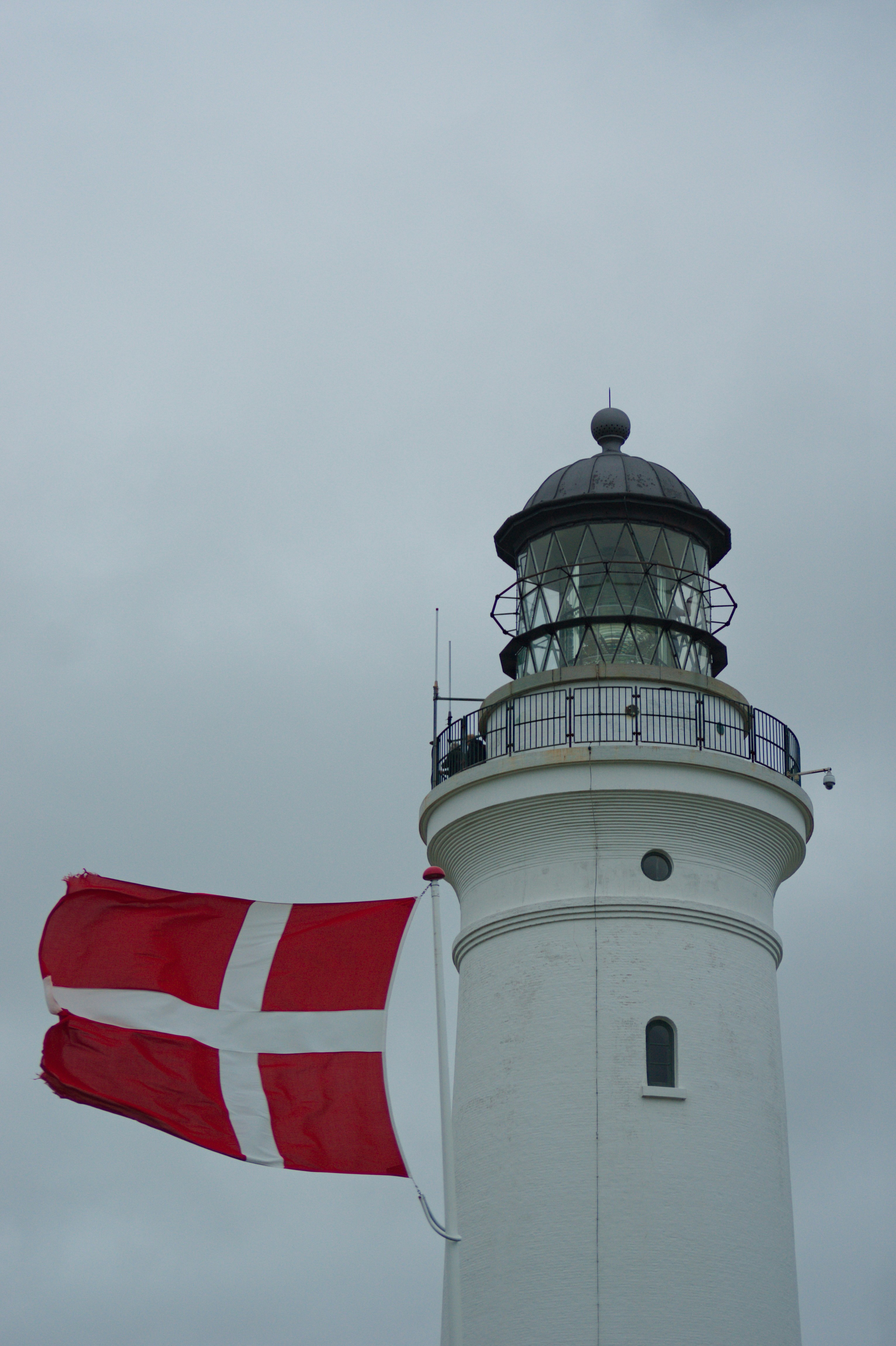 A tall lighthouse stands against a cloudy sky, with a vibrant Danish flag flying prominently. The structure embodies maritime heritage and resilience.