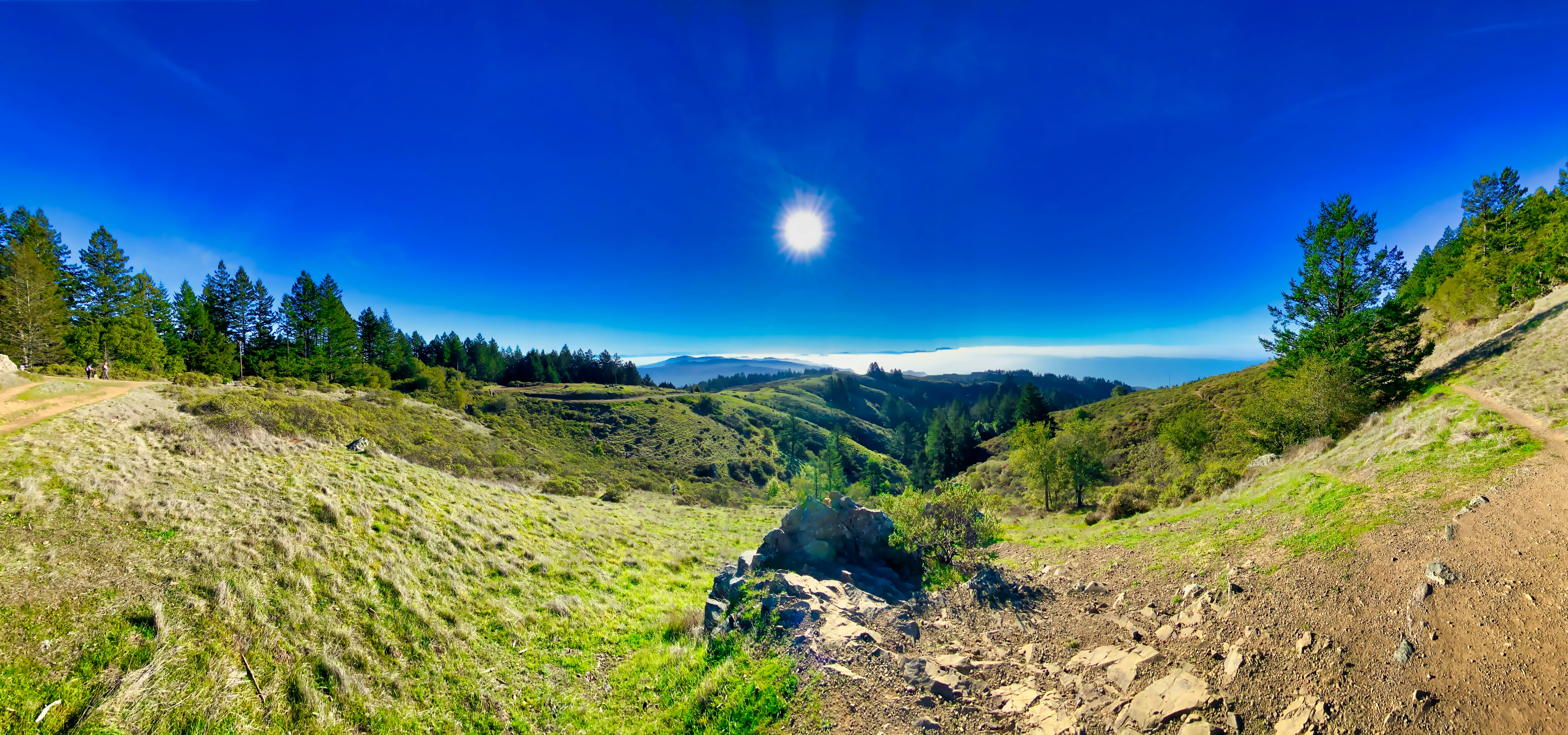 Vast green valley under a clear blue sky, with the sun shining brightly above and rolling hills in the background.