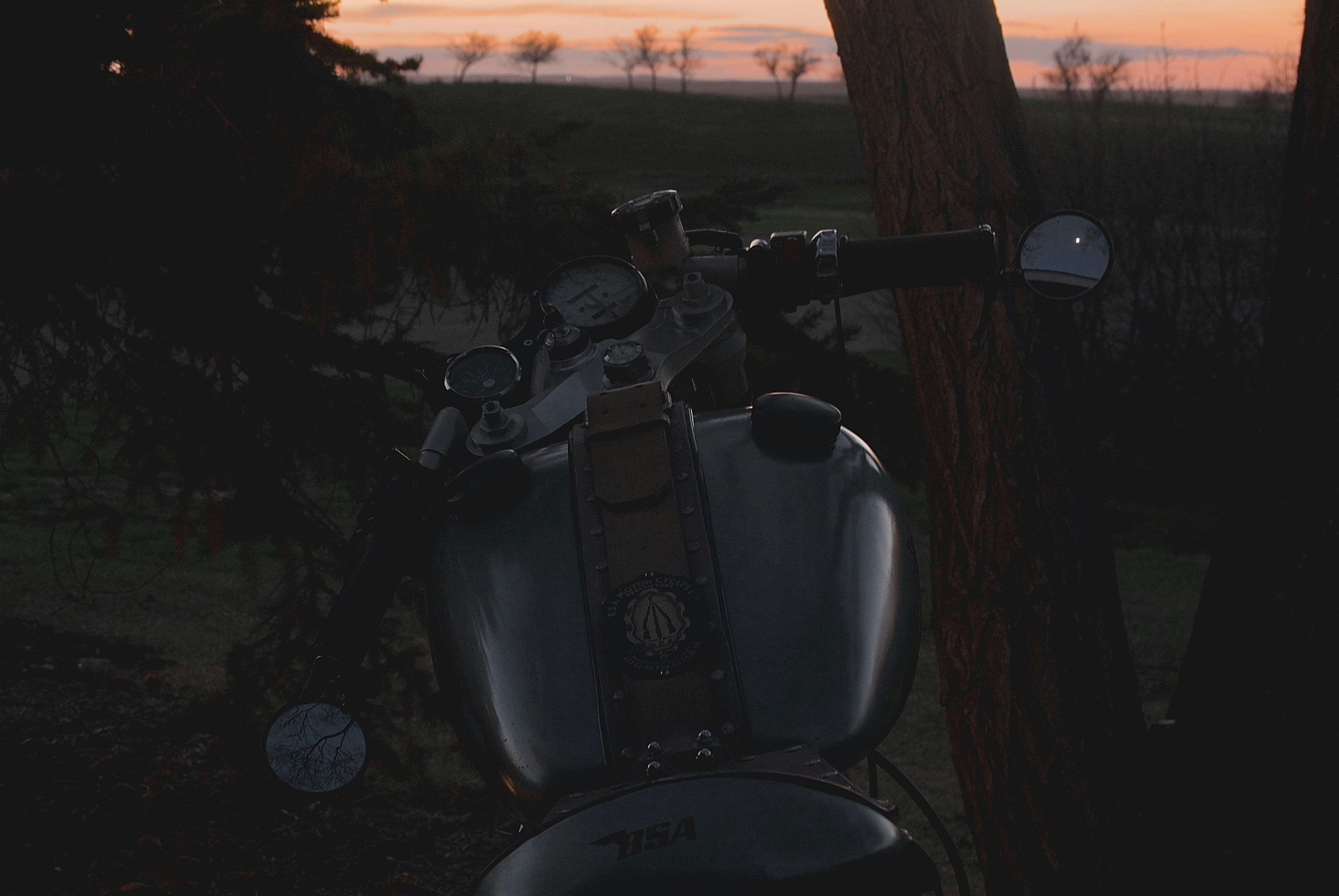 Vintage motorcycle silhouette against a fading sunset, with a rugged tank and handlebars dominating the foreground. The calm horizon and warm sky create a moody, nostalgic atmosphere.
