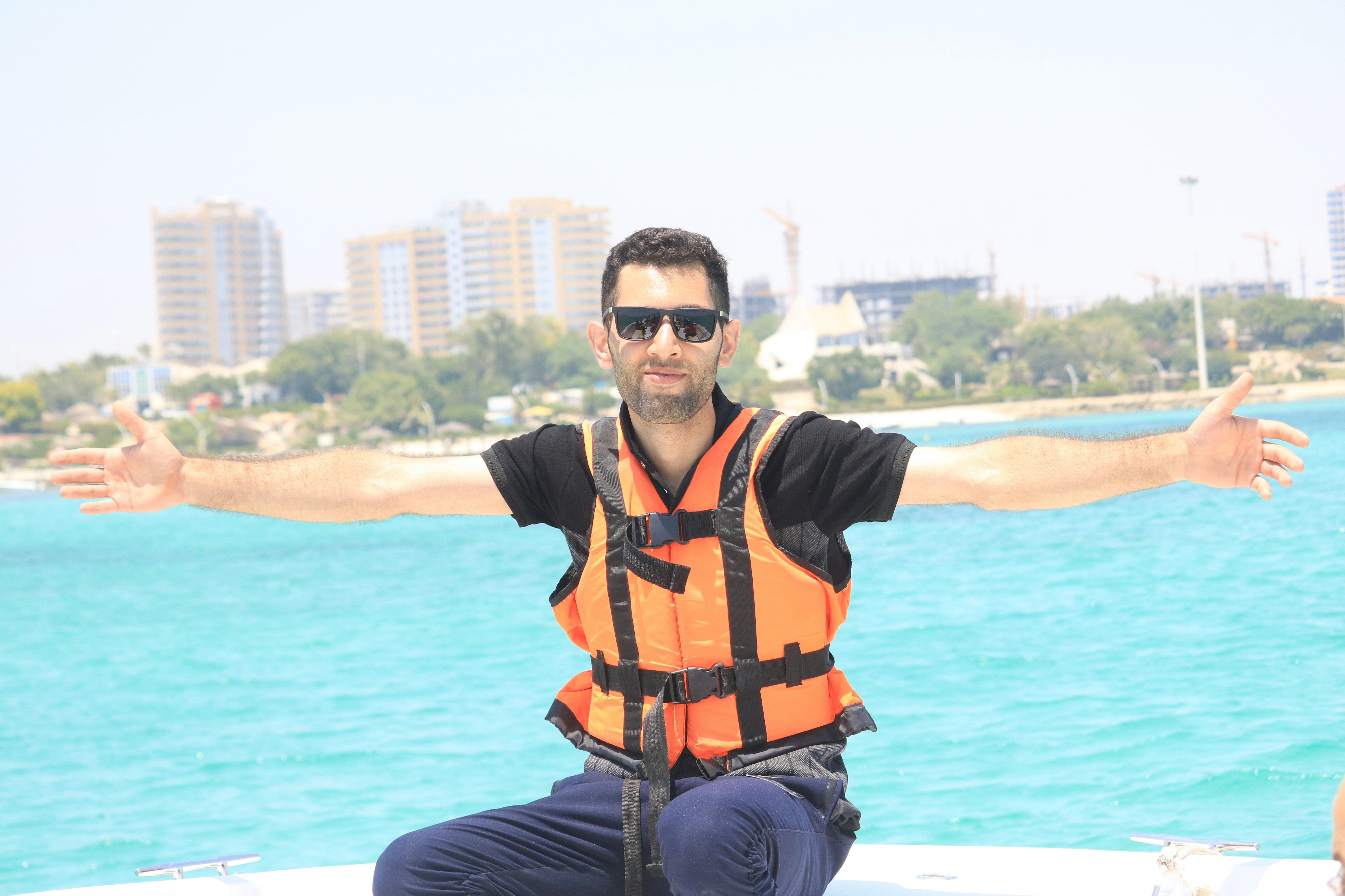 man in orange and black life vest and black sunglasses sitting on the beach during daytime