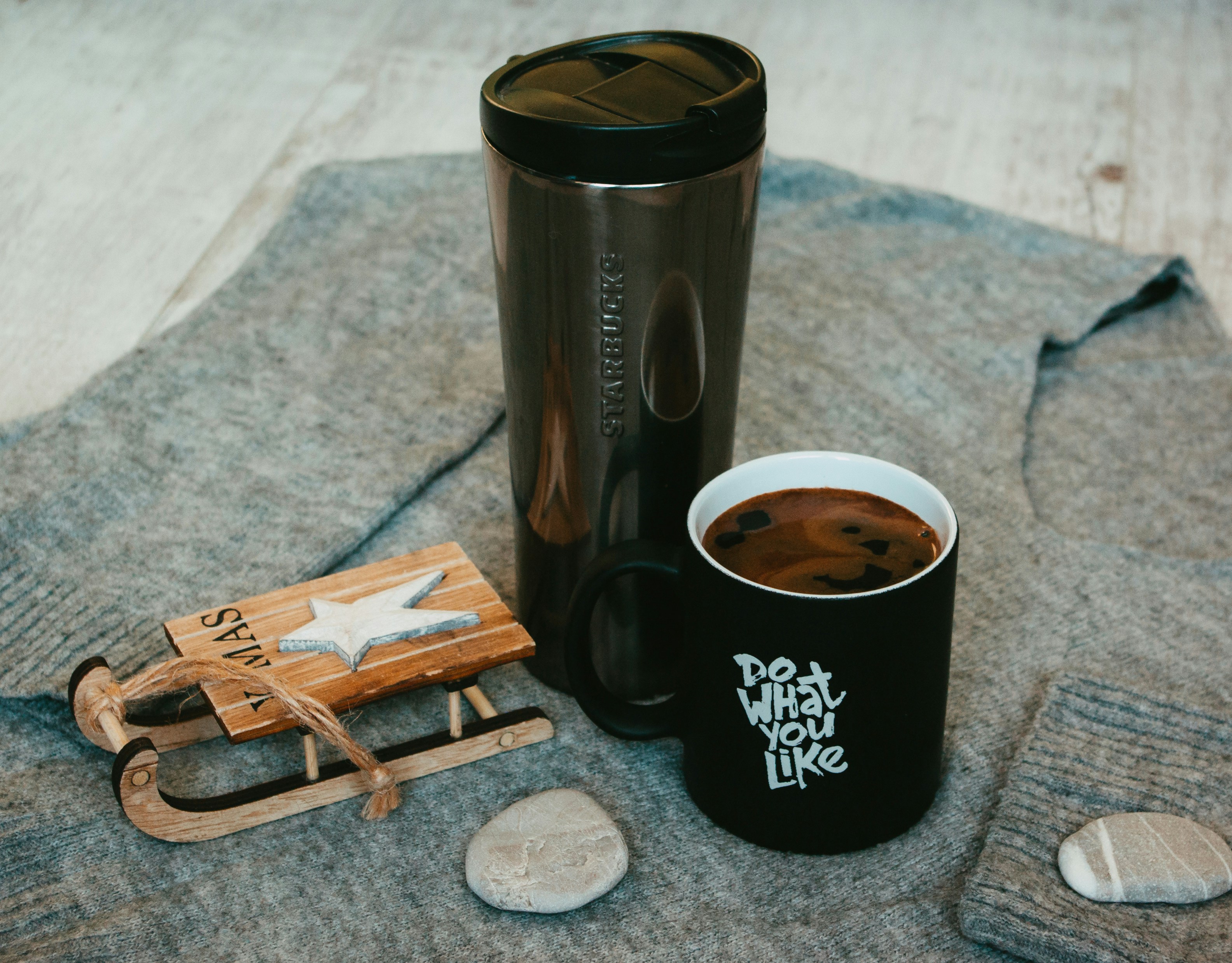 A black mug with an inspiring message sits beside a stainless steel travel tumbler and wooden sled on a soft gray sweater. Smooth stones add a touch of nature to the setting.