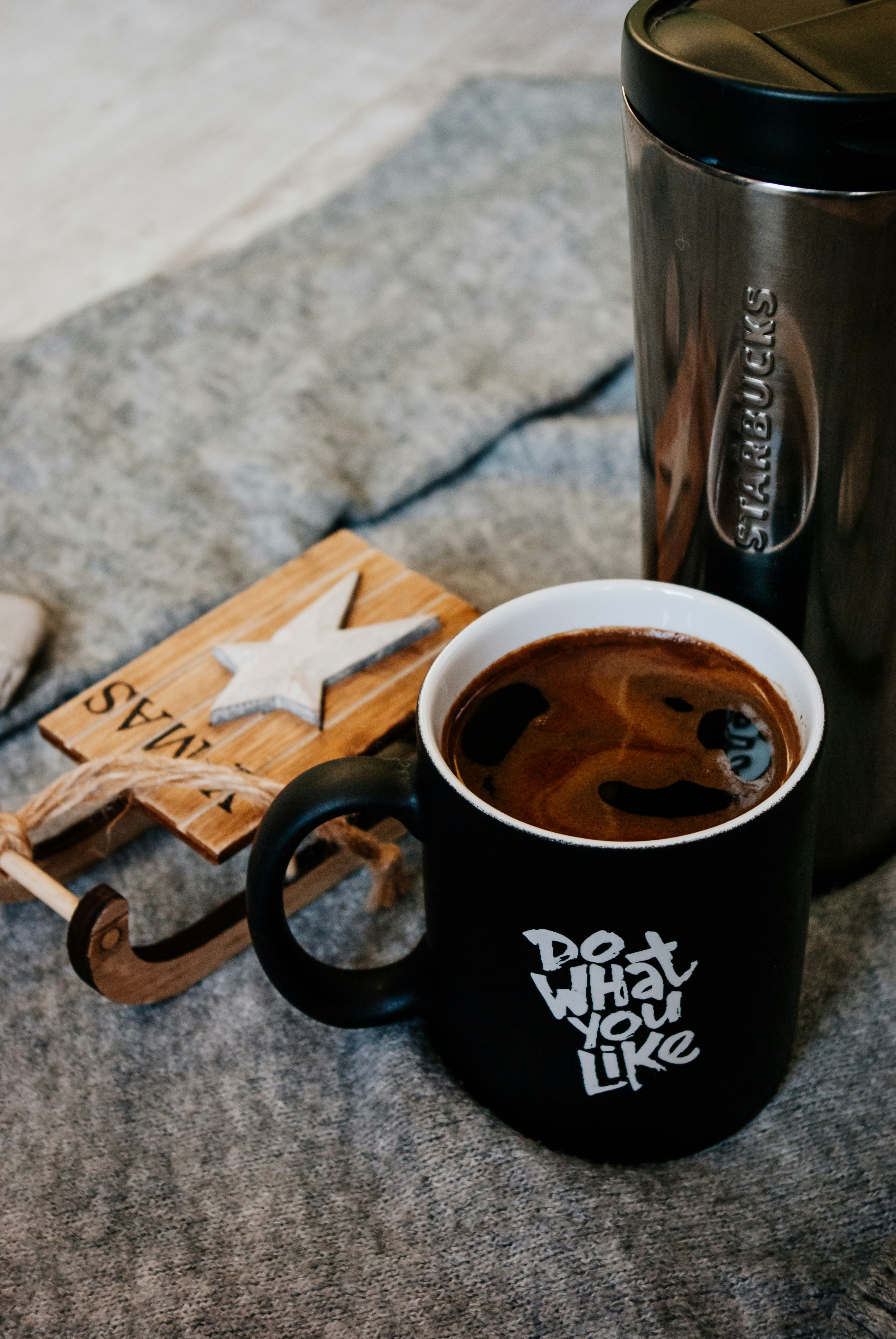 Close-up of a black mug reading 'Do What You Like' brimming with coffee, set on a textured fabric beside a stainless steel Starbucks-branded tumbler and a wooden star ornament.
