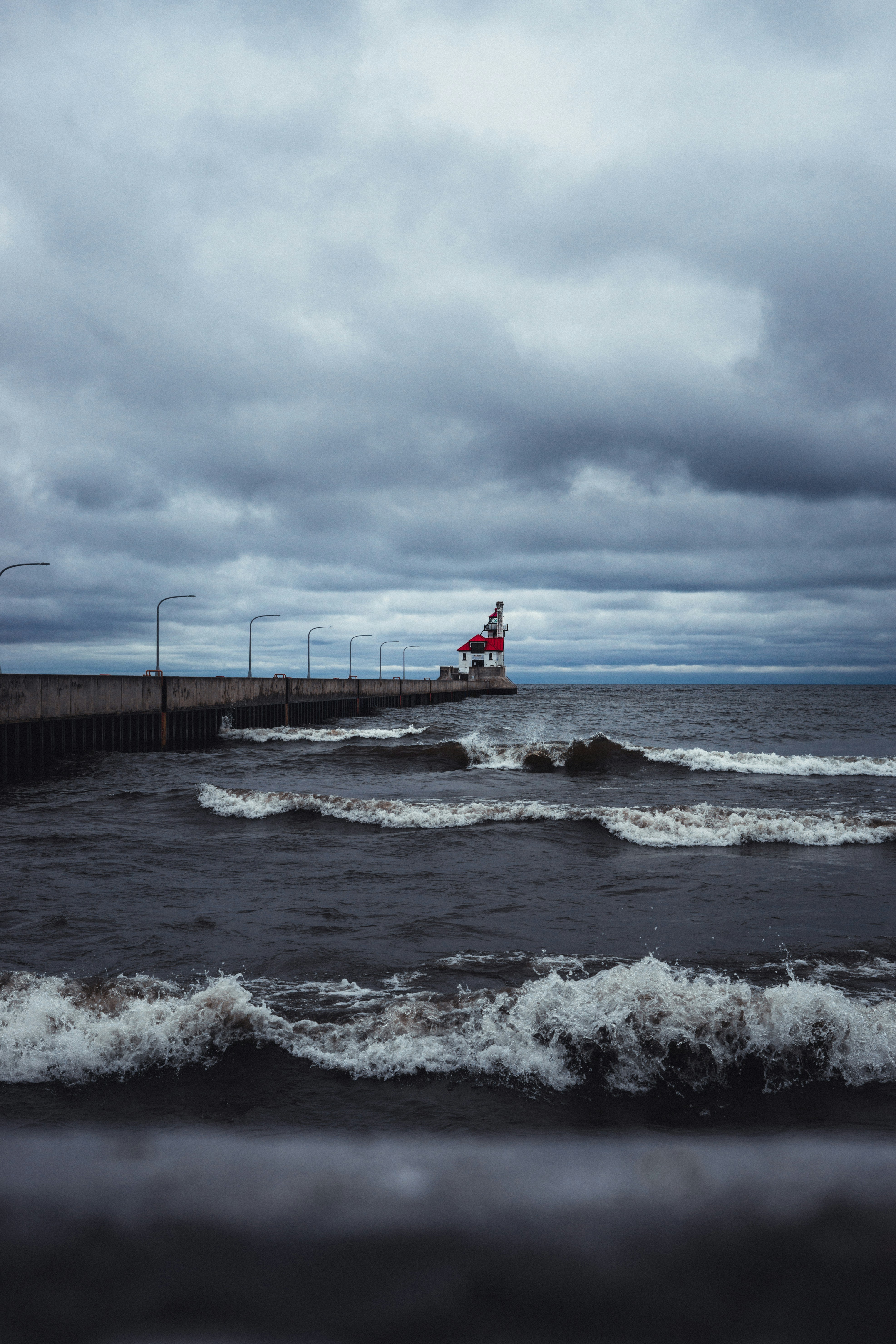 Waves crash against a pier leading to a red-roofed lighthouse under a cloudy sky.