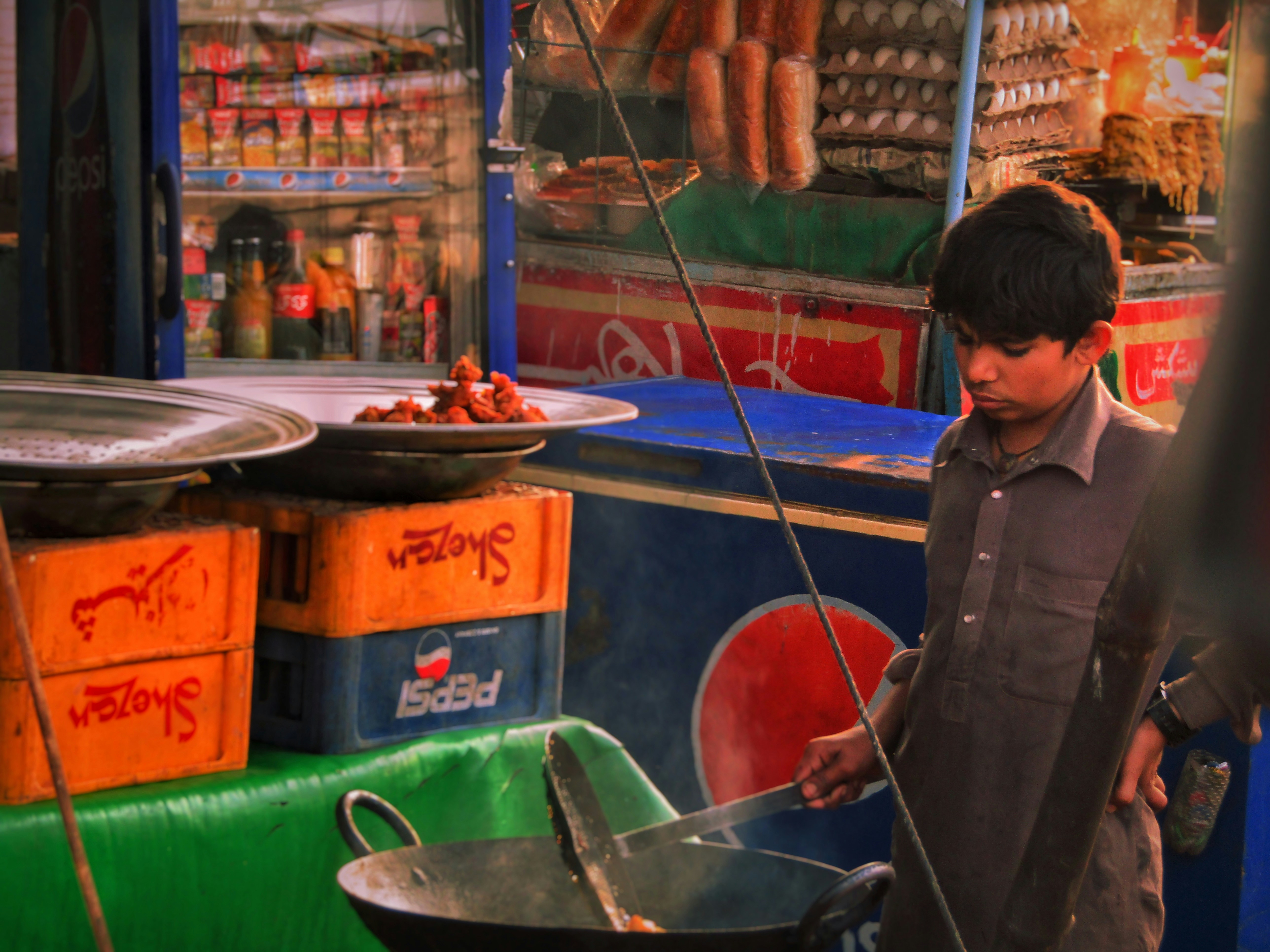 Young vendor preparing food at a colorful street stall, surrounded by stacked crates and vibrant displays.