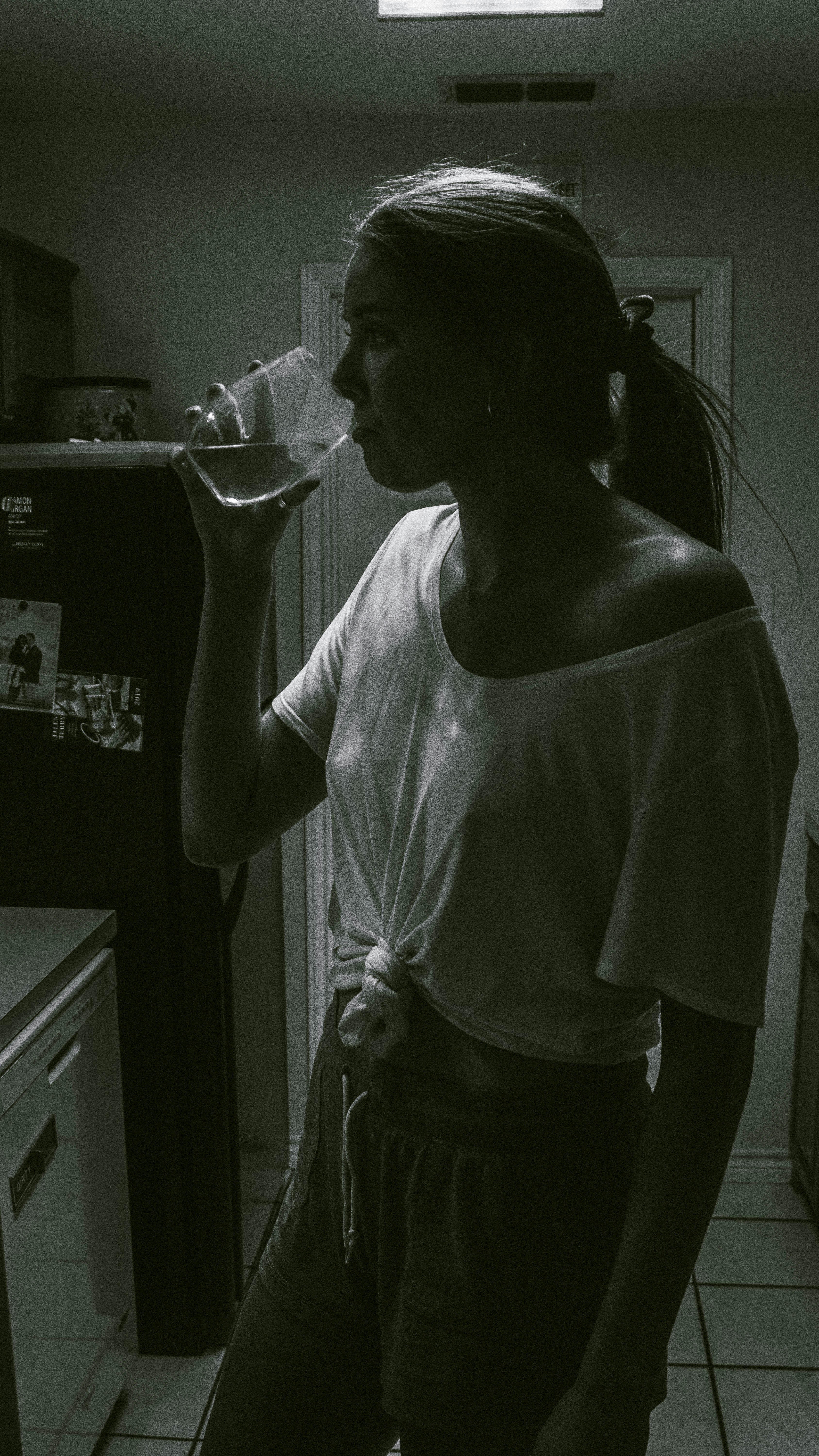 A young woman sipping water in a dimly lit kitchen, capturing a moment of introspection and calm. The scene is enriched by soft shadows and a minimalist aesthetic.