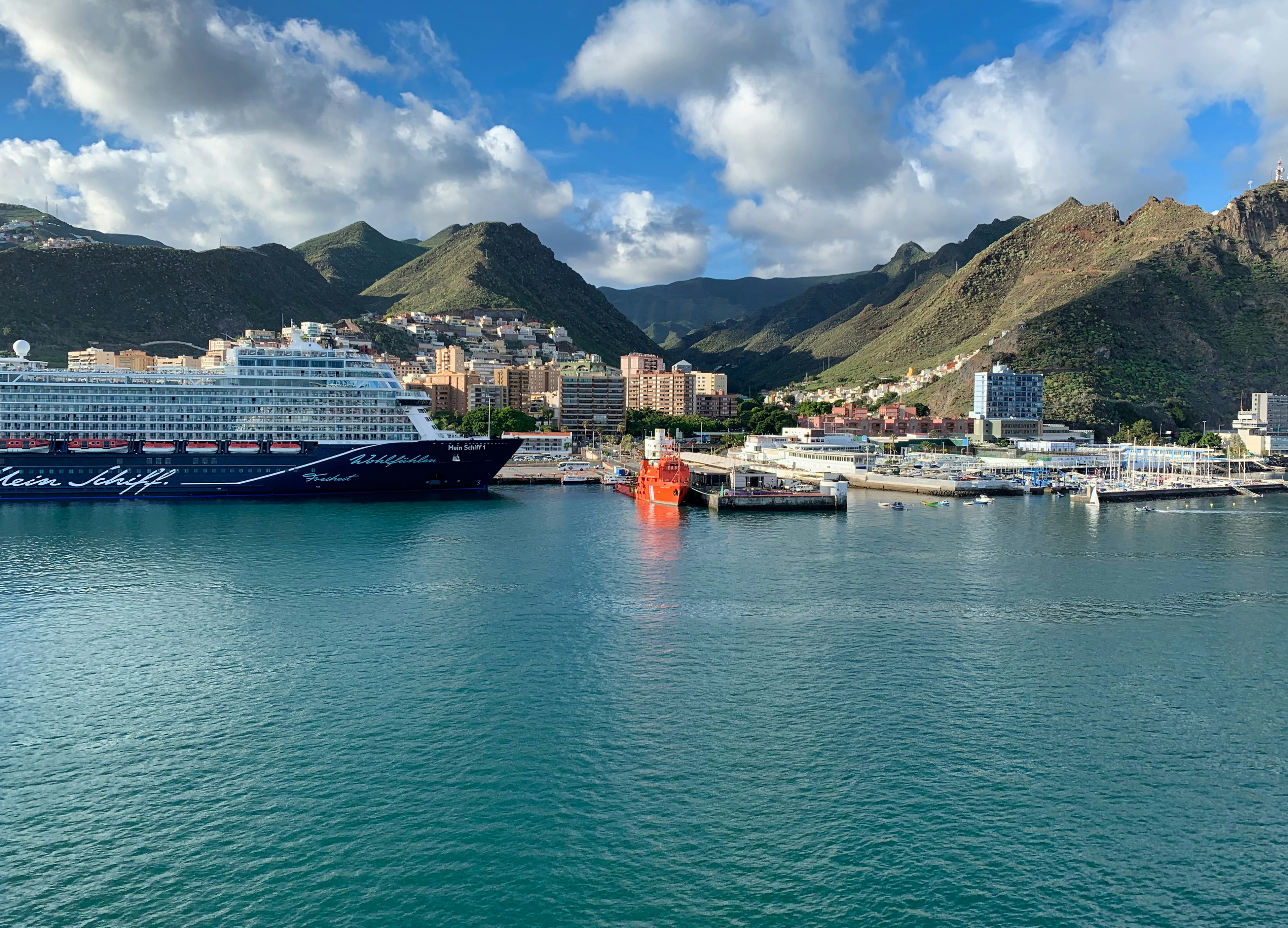 Large cruise ship docked in a tranquil harbor surrounded by mountains and urban architecture. Bright blue waters reflect the sky.