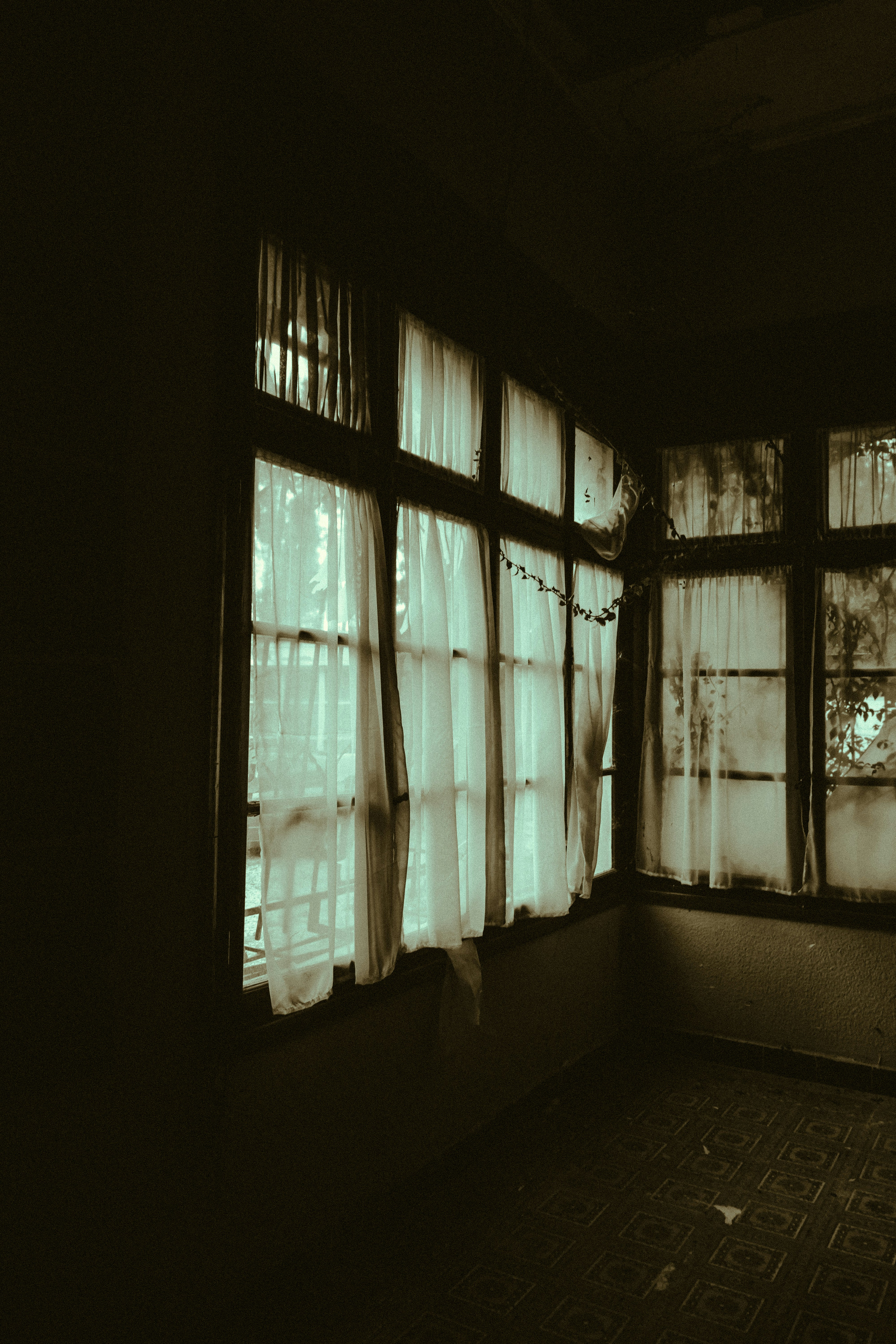 Sunlight filtering through aged, sheer curtains in a dimly lit room, casting soft shadows on the floor.