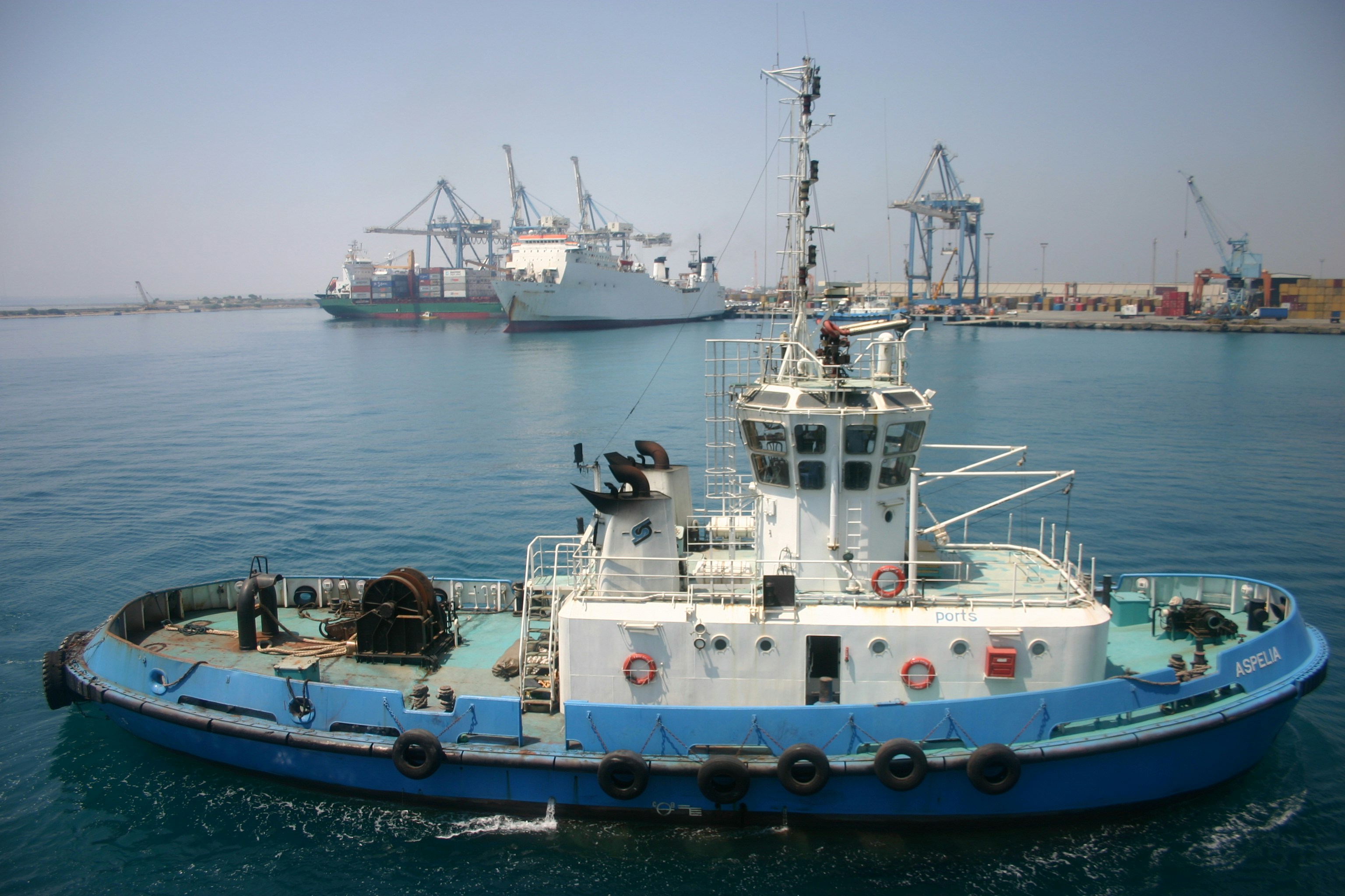 Blue and white tugboat navigating a calm harbor with cranes and ships in the background.