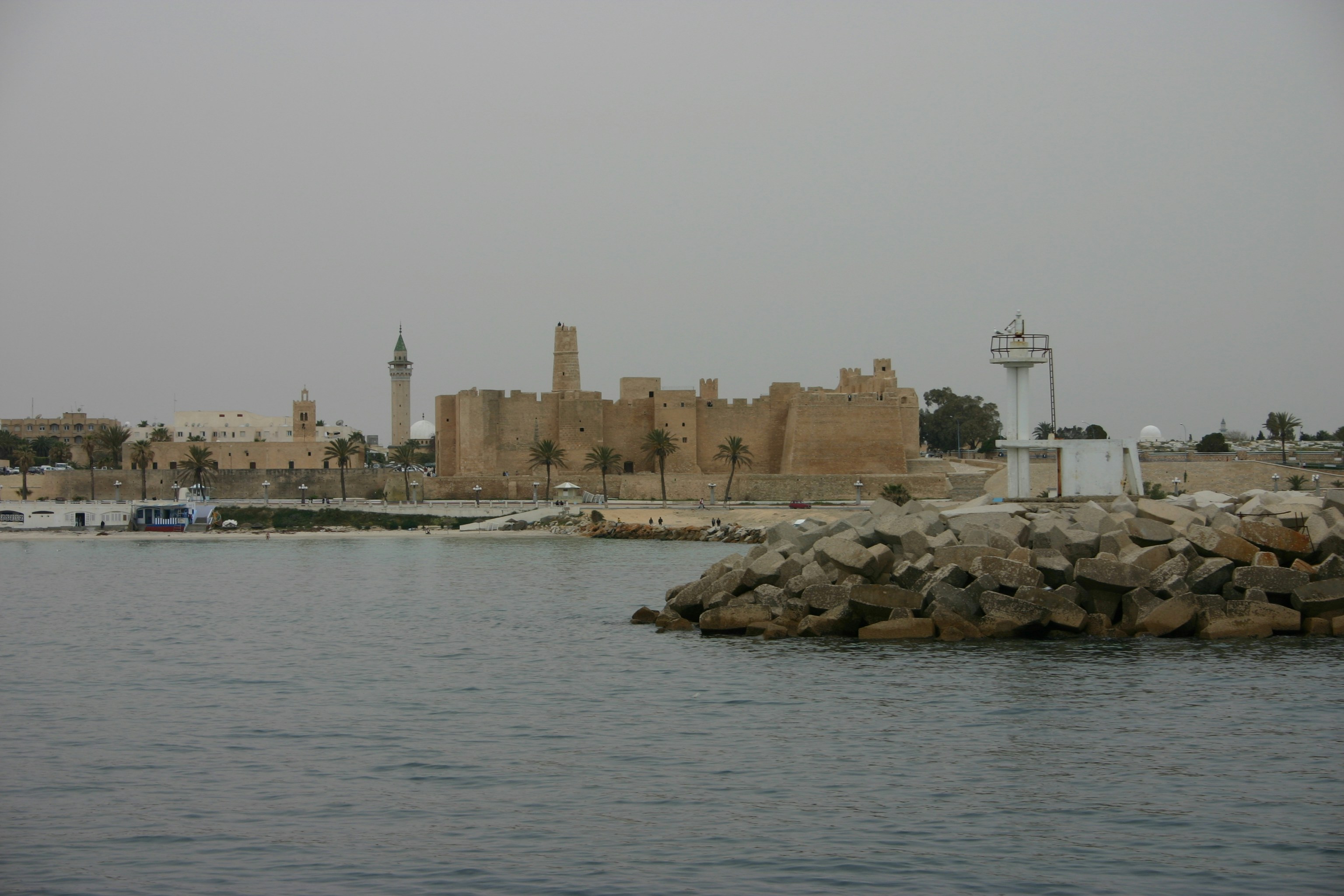 Ancient fortress stands resilient against the coastal backdrop, with a lighthouse and boats nearby. A serene scene capturing the blend of nature and architecture.