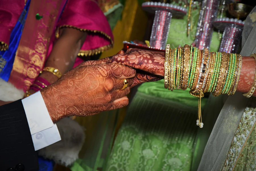 Henna-covered hands exchange a ring beside stacks of gold and green bangles during a wedding ceremony. Bright pink and green fabrics frame the intimate moment.