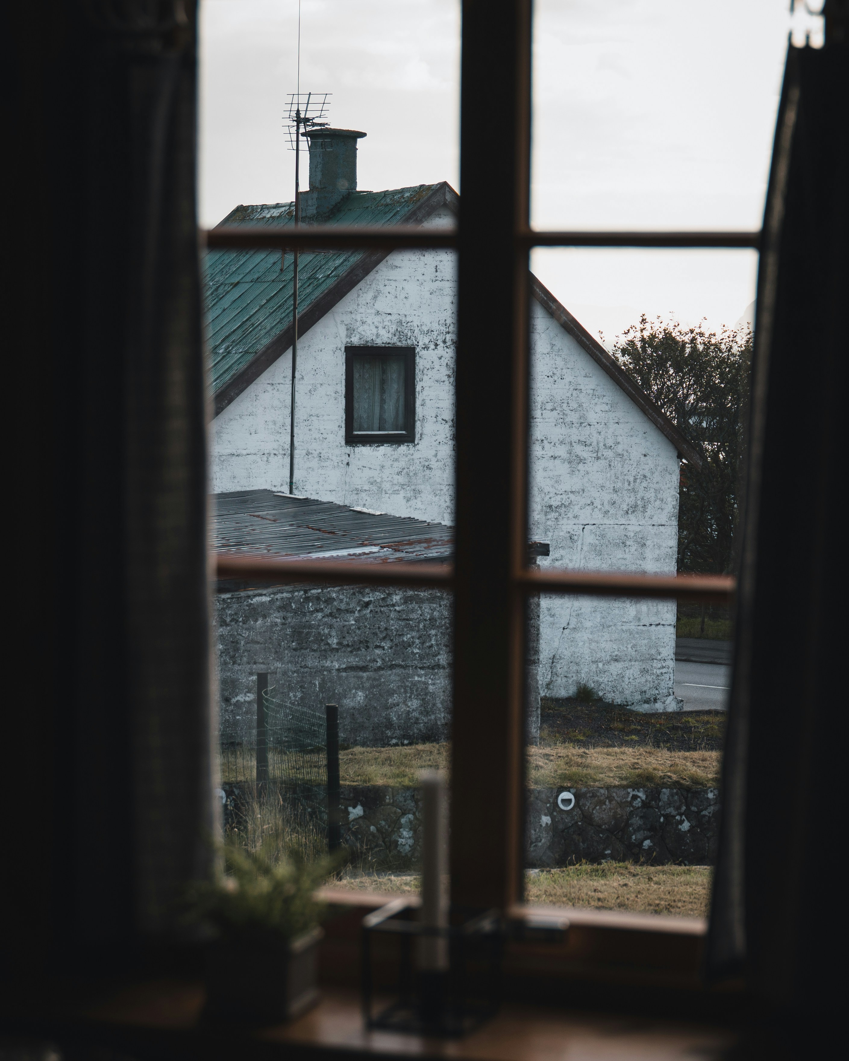 View of a rustic white house with a green roof framed by a window, showcasing the serene countryside beyond.