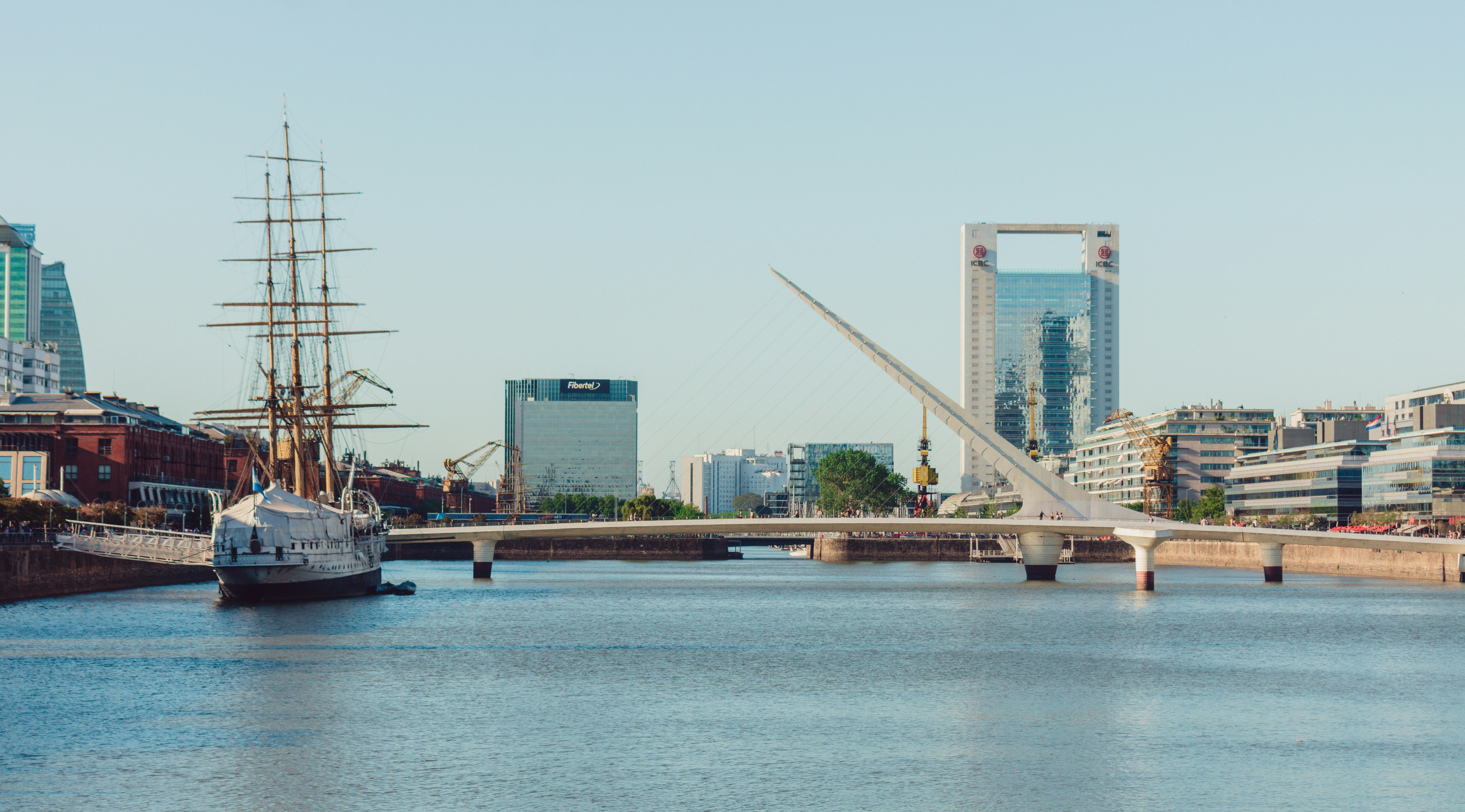 Historic sailing ship moored beside a modern cable-stayed bridge over a tranquil urban river.