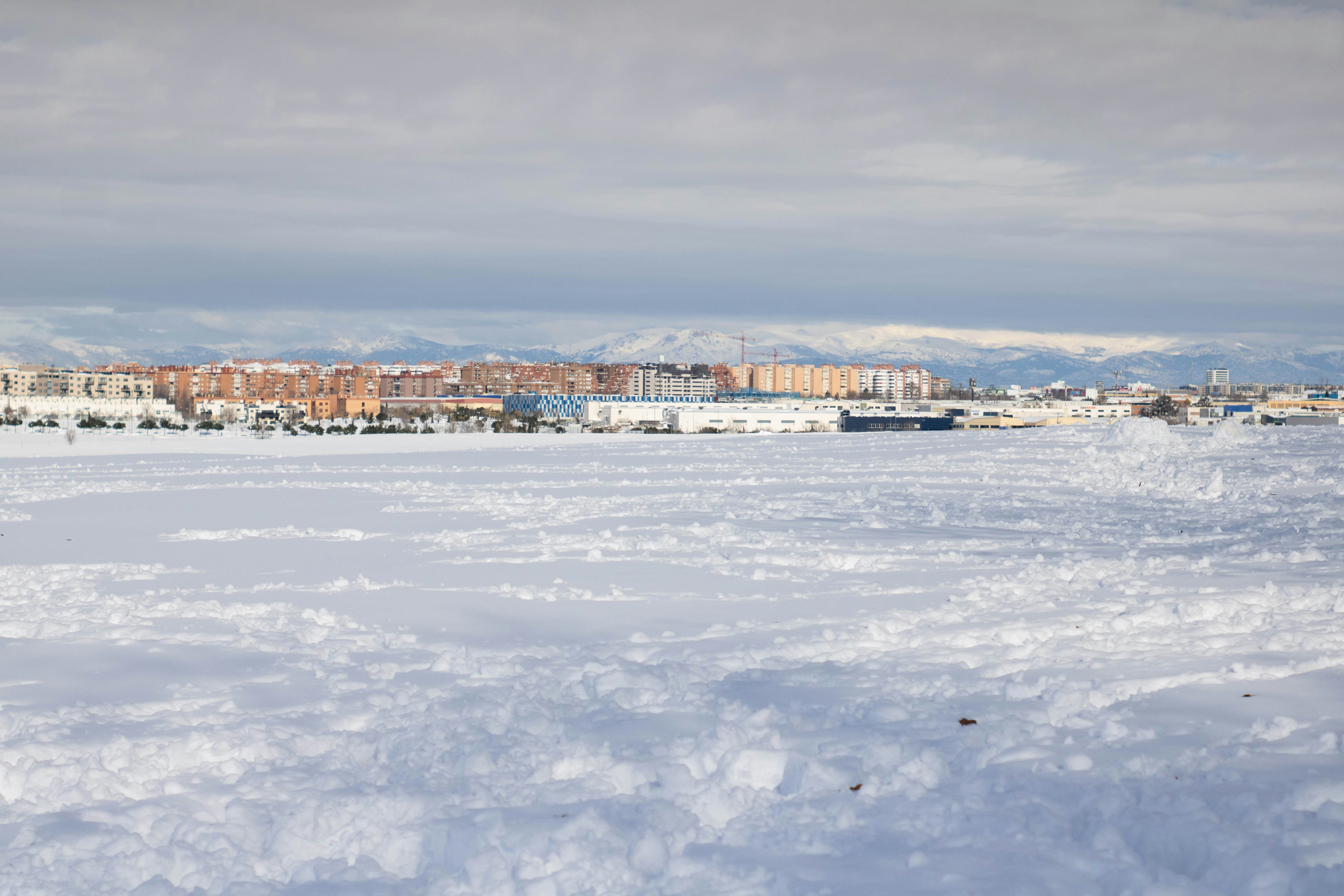 A vast expanse of snow blankets the ground, with a city skyline visible in the background under a cloudy sky.