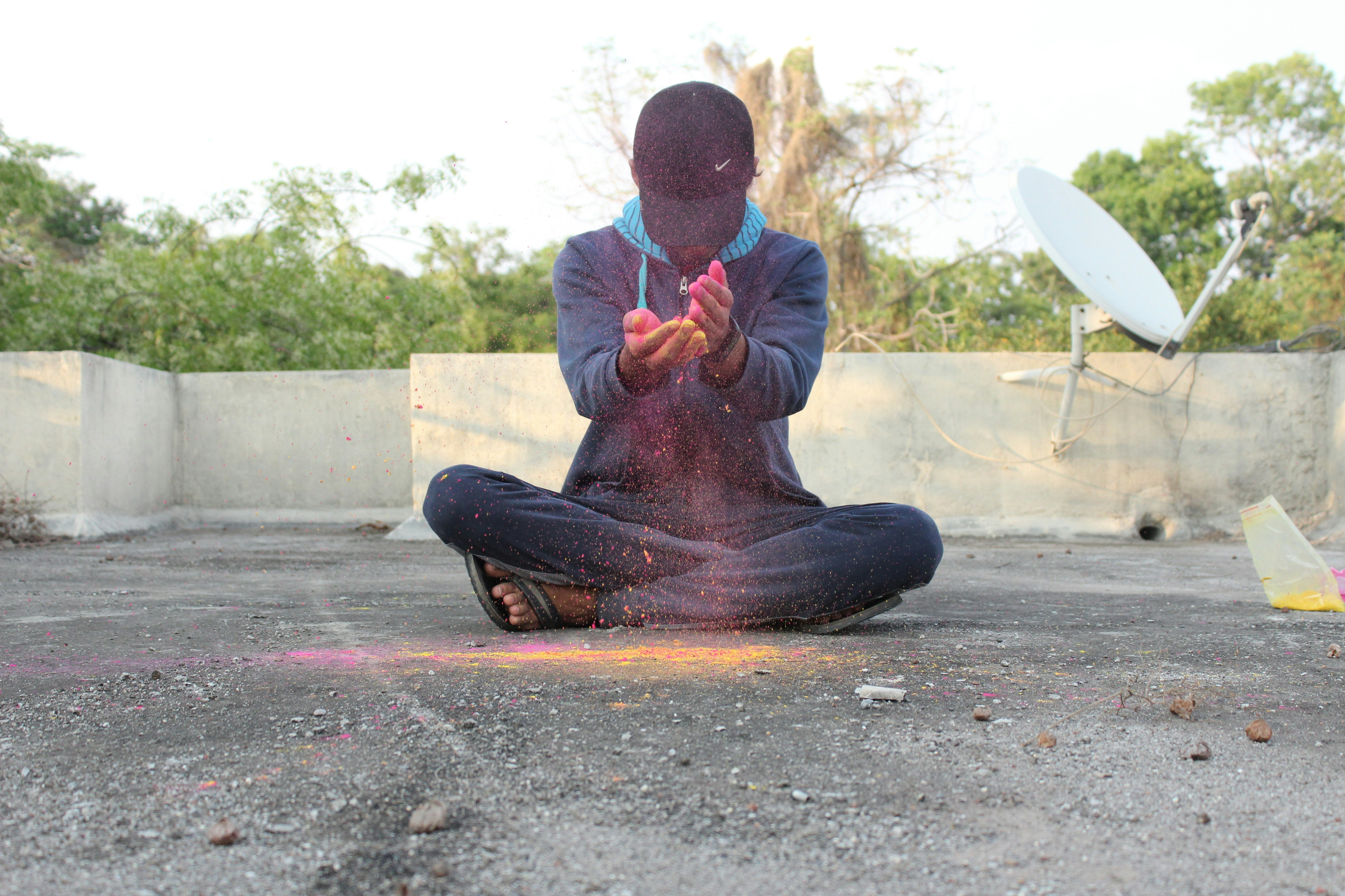 Person in blue attire sitting cross-legged on a rooftop, releasing vibrant powders into the air.