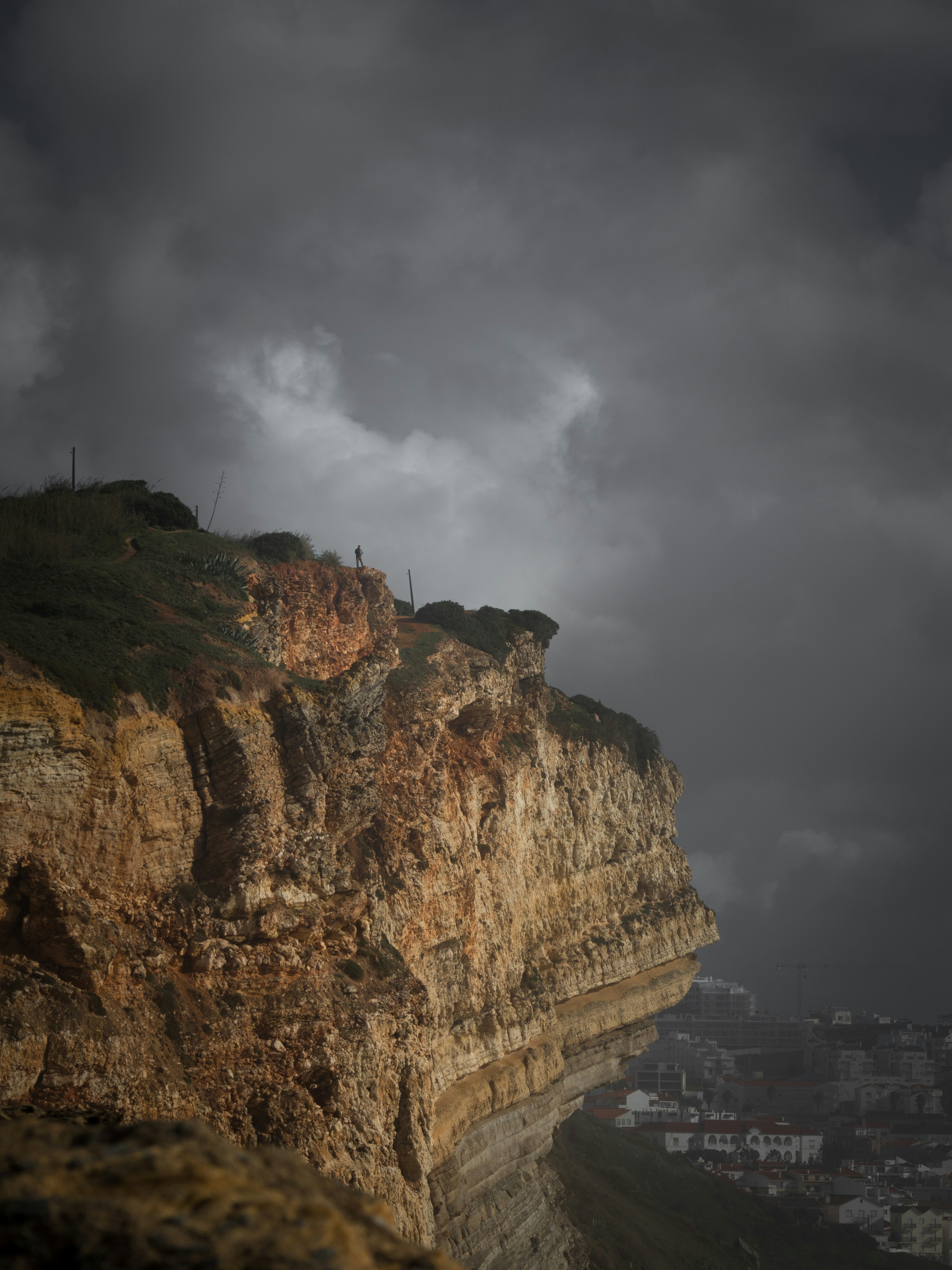 A rugged cliff rising dramatically under a moody sky, with a solitary figure standing at the precipice, contemplating the vast landscape below.