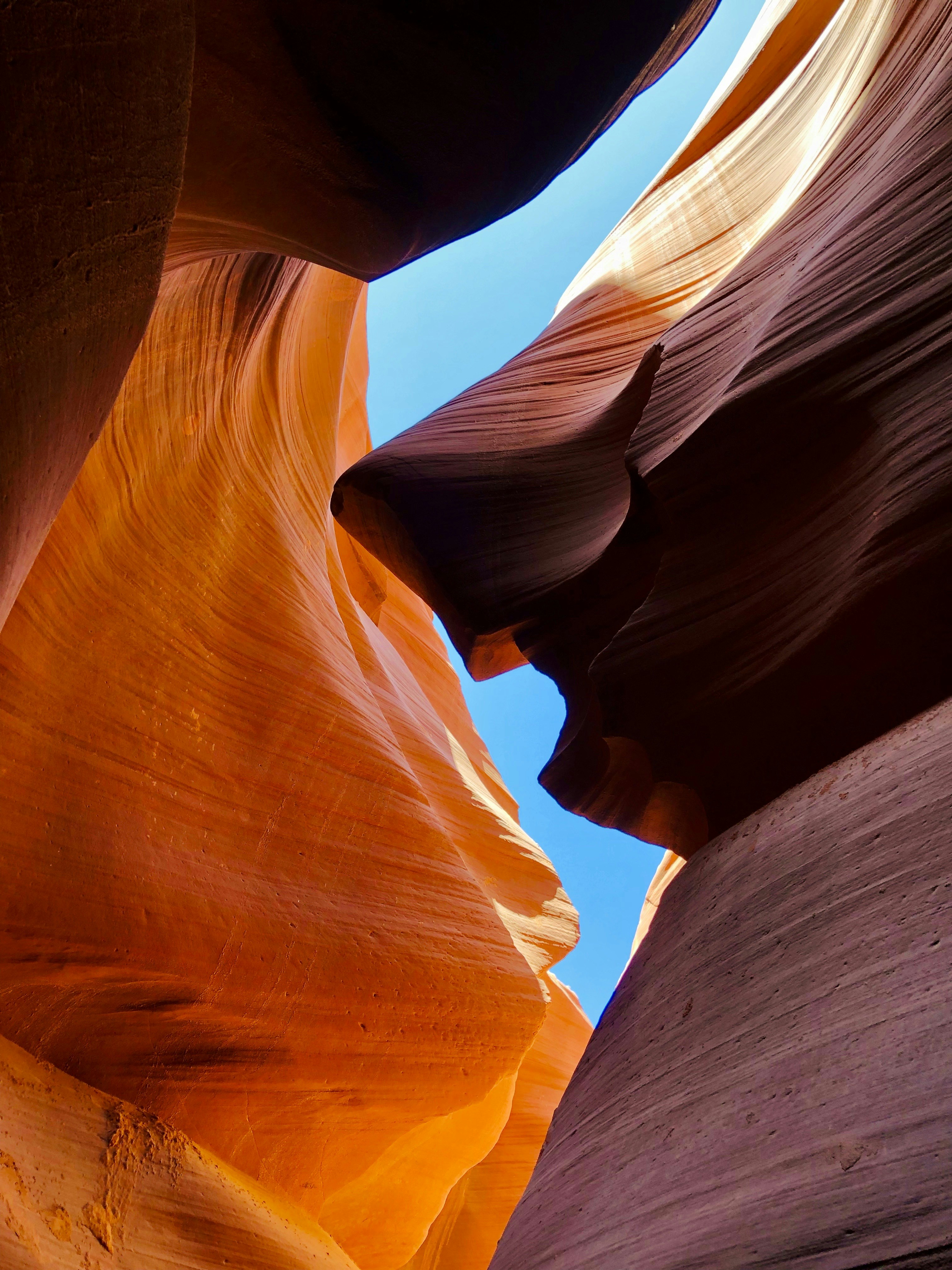 Vibrant orange and brown sandstone formations converge towards a bright blue sky in a narrow canyon. The interplay of light and shadow highlights the natural curves and textures of the rock walls.