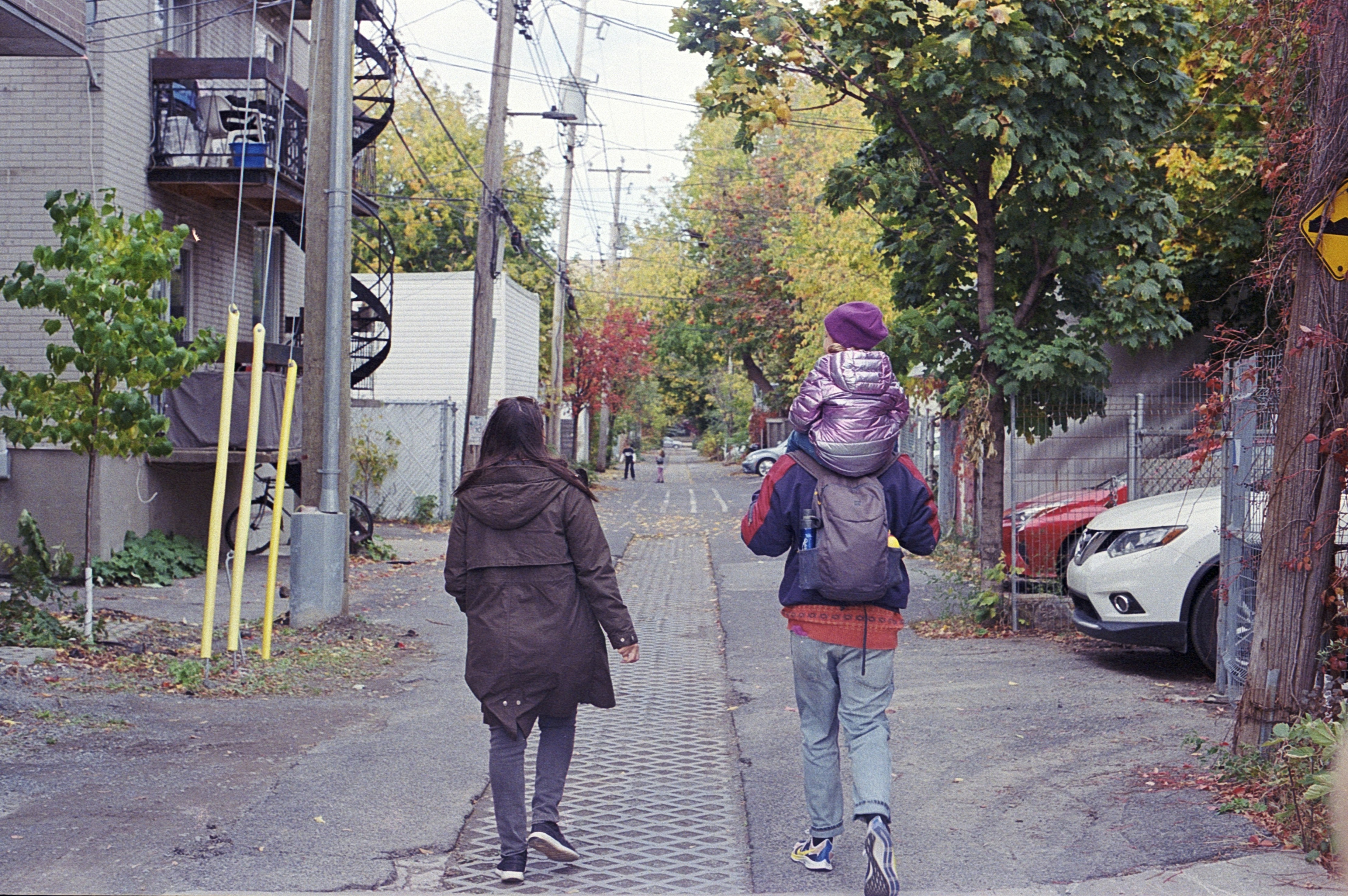 Photograph of two figures walking away along a narrow street lined with trees and parked cars; a parent carries a child on shoulders beneath autumn light.