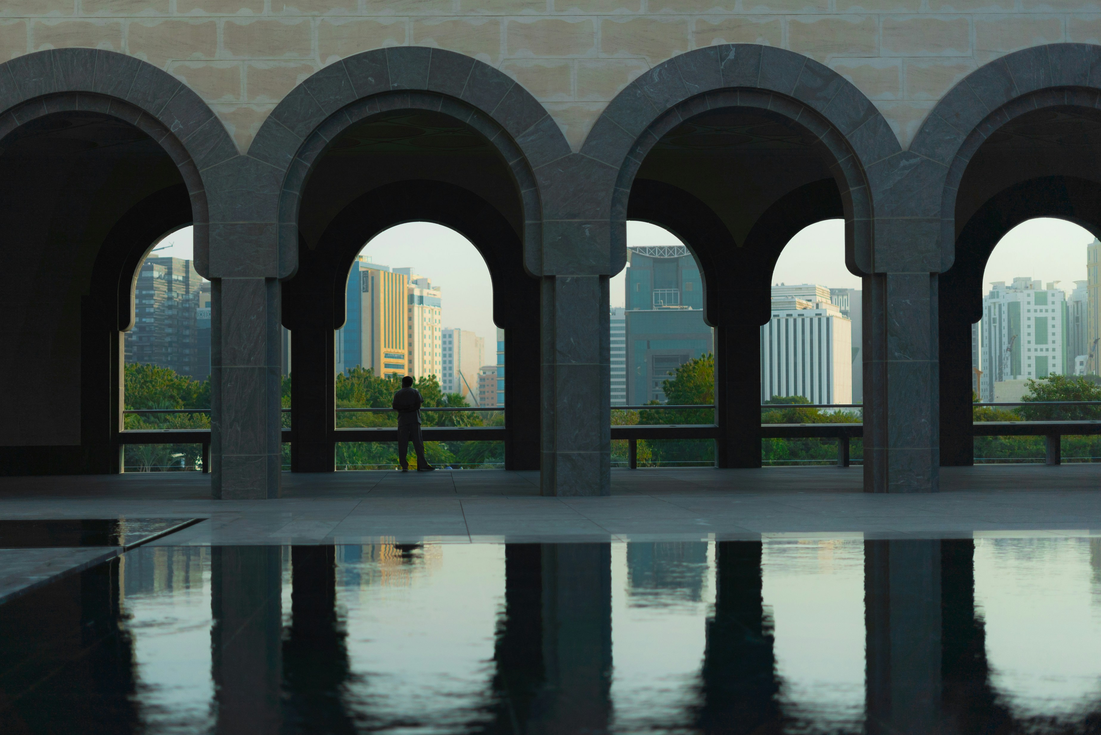 Silhouette of a person framed by arched windows overlooking a cityscape with mirrored reflections on a calm surface.