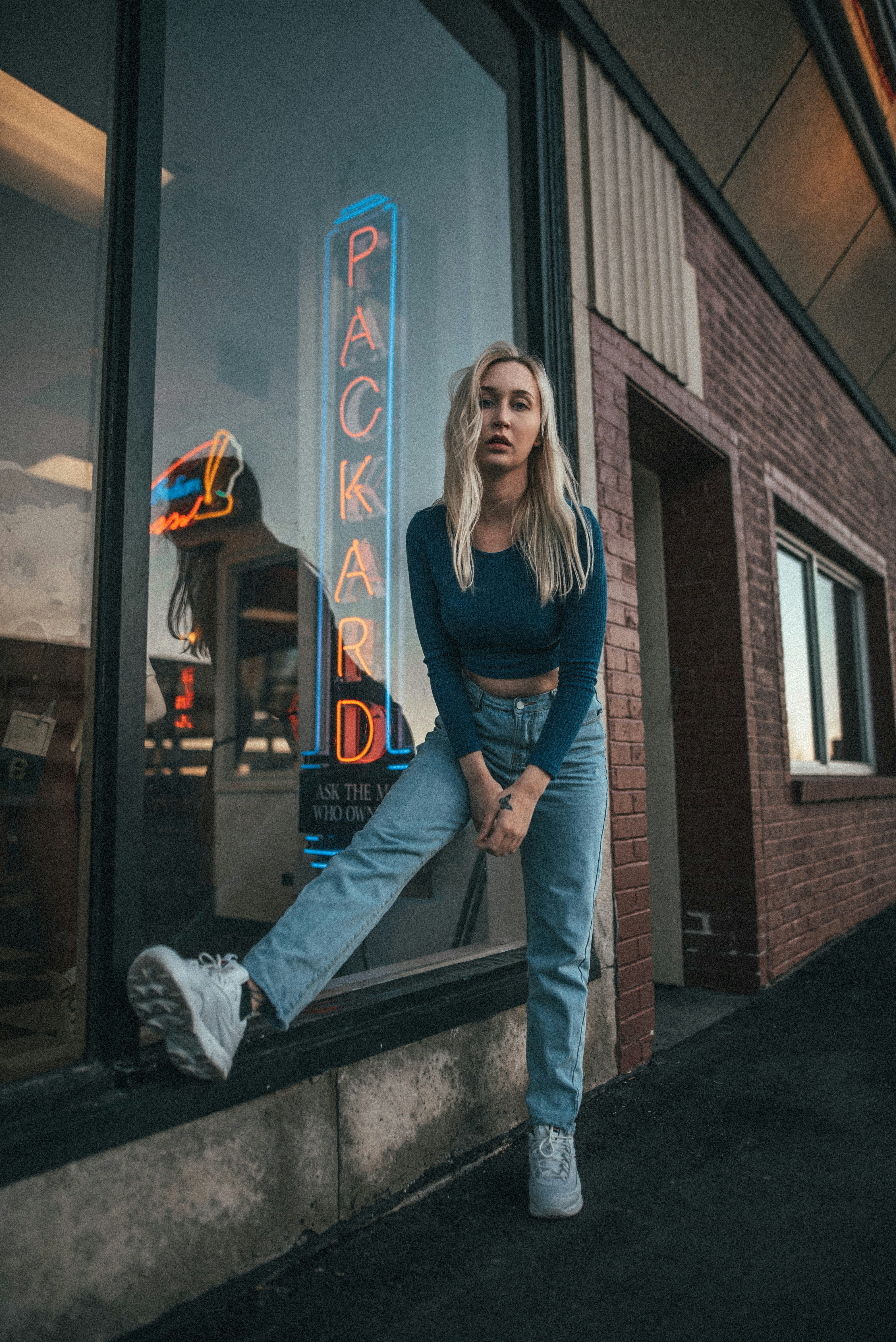 Young woman striking a pose in front of a neon sign, embodying urban fashion against a backdrop of brick and glass.