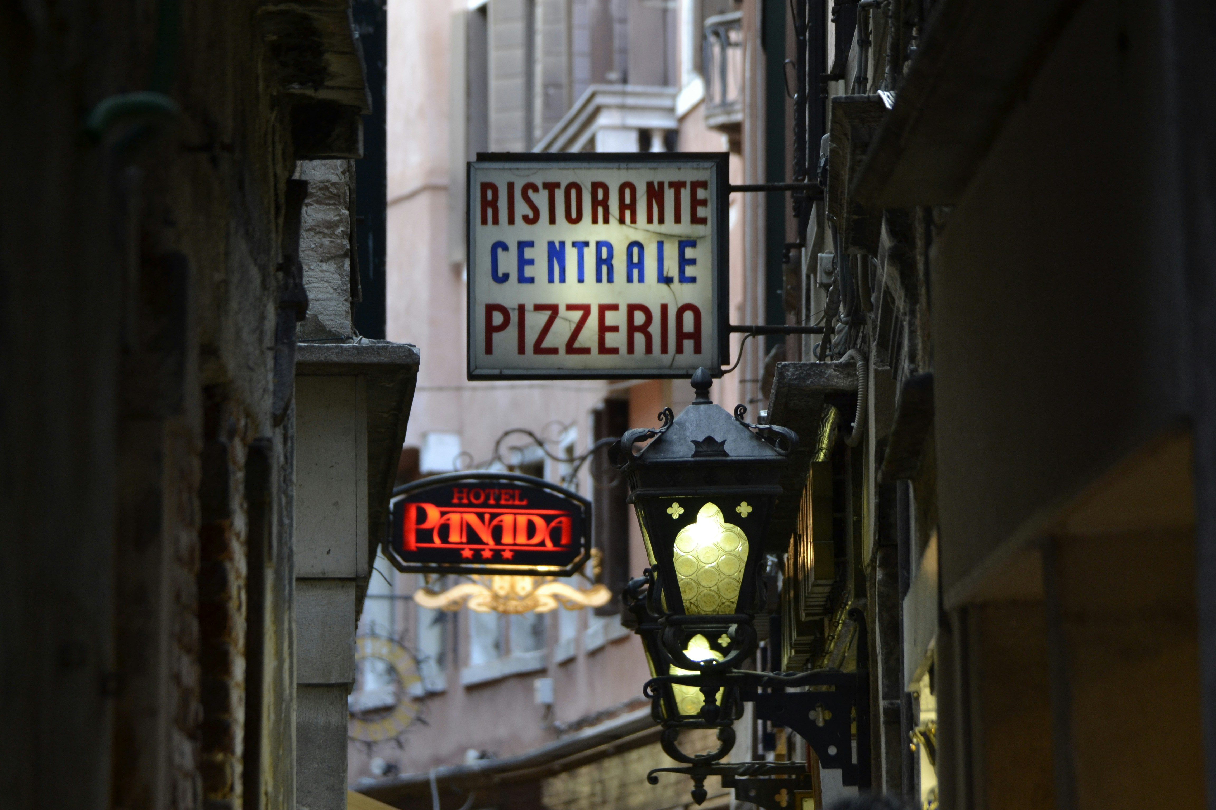 Night-time Italian alley bathed in warm lantern light and neon signage. The scene centers on the RISTORANTE CENTRALE PIZZERIA sign amid narrow stone walls.