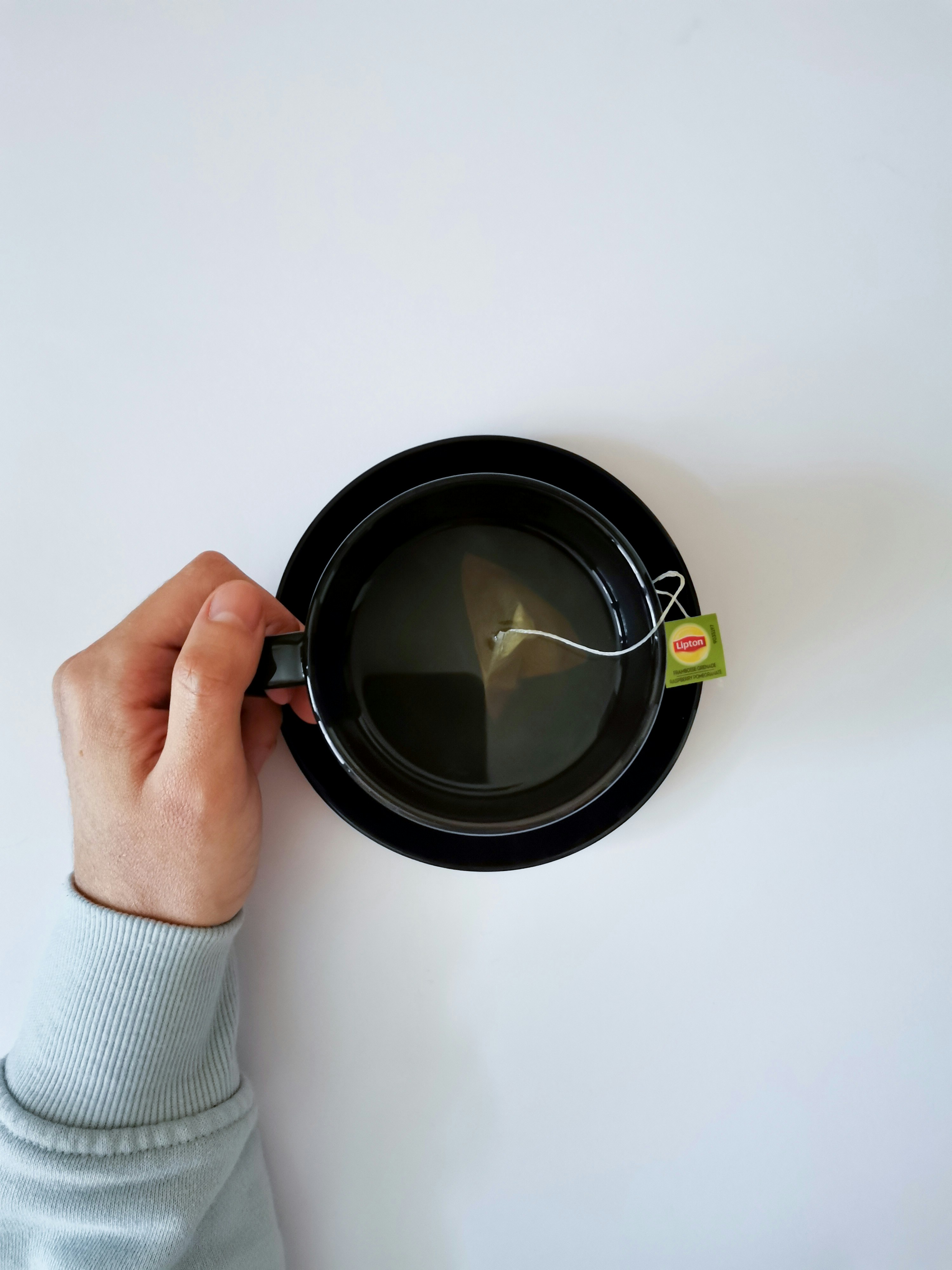 Hand holding a black cup of tea with a teabag tag resting on the rim, set against a minimalistic white background.
