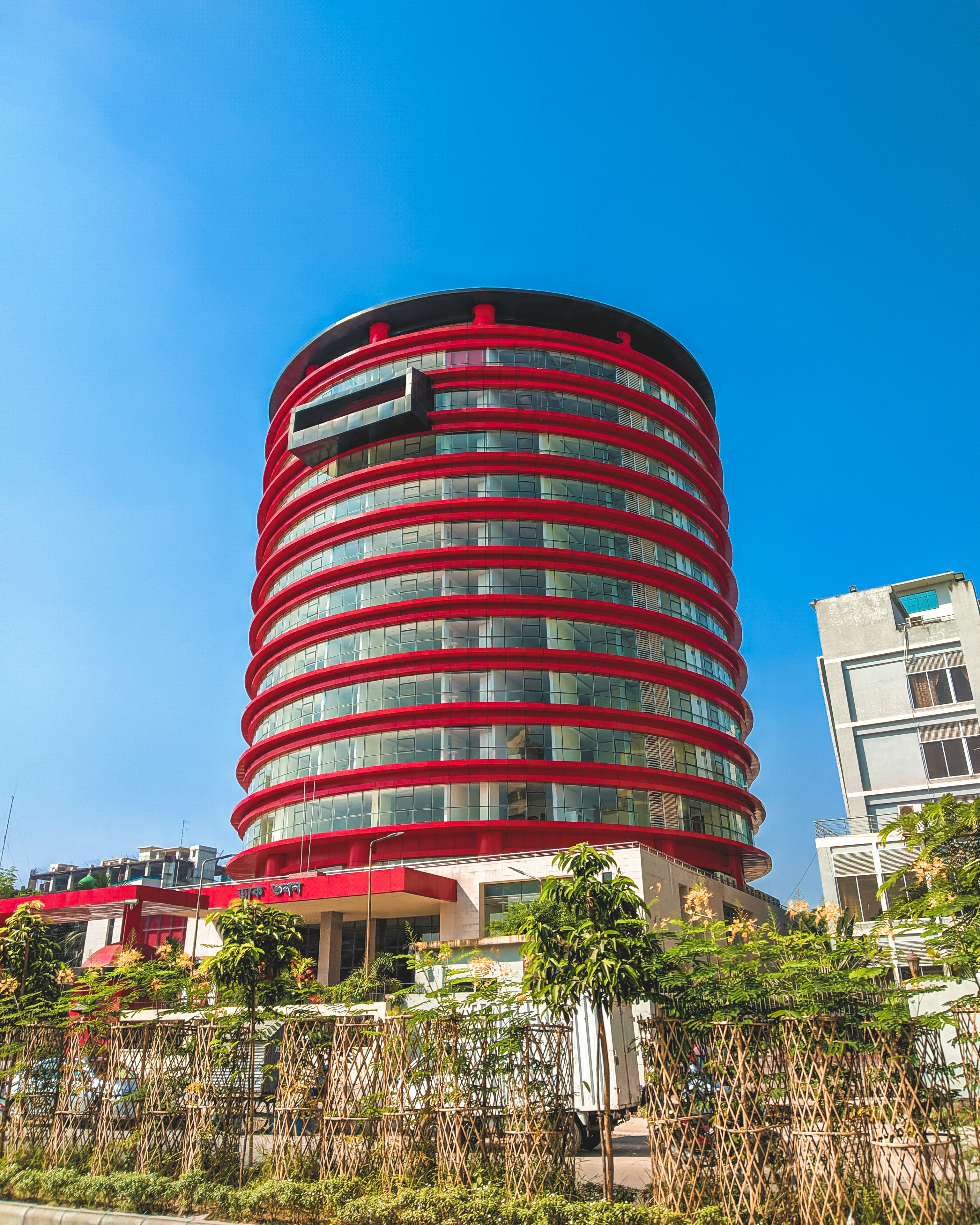 Vibrant red circular building against a clear blue sky, showcasing contemporary architectural design with lush greenery in the foreground.